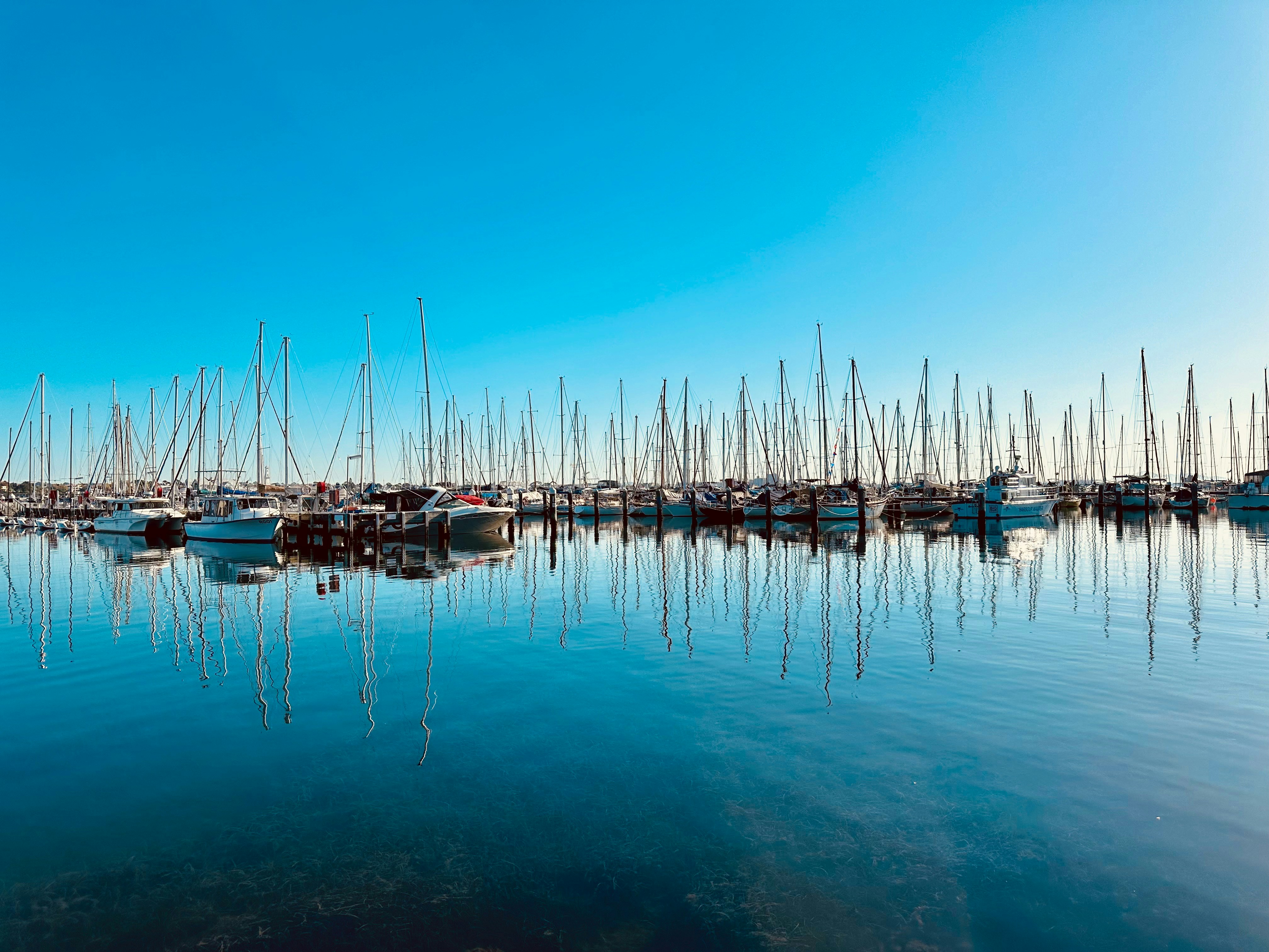 Sailboats reflected in calm harbor waters under a bright blue sky.