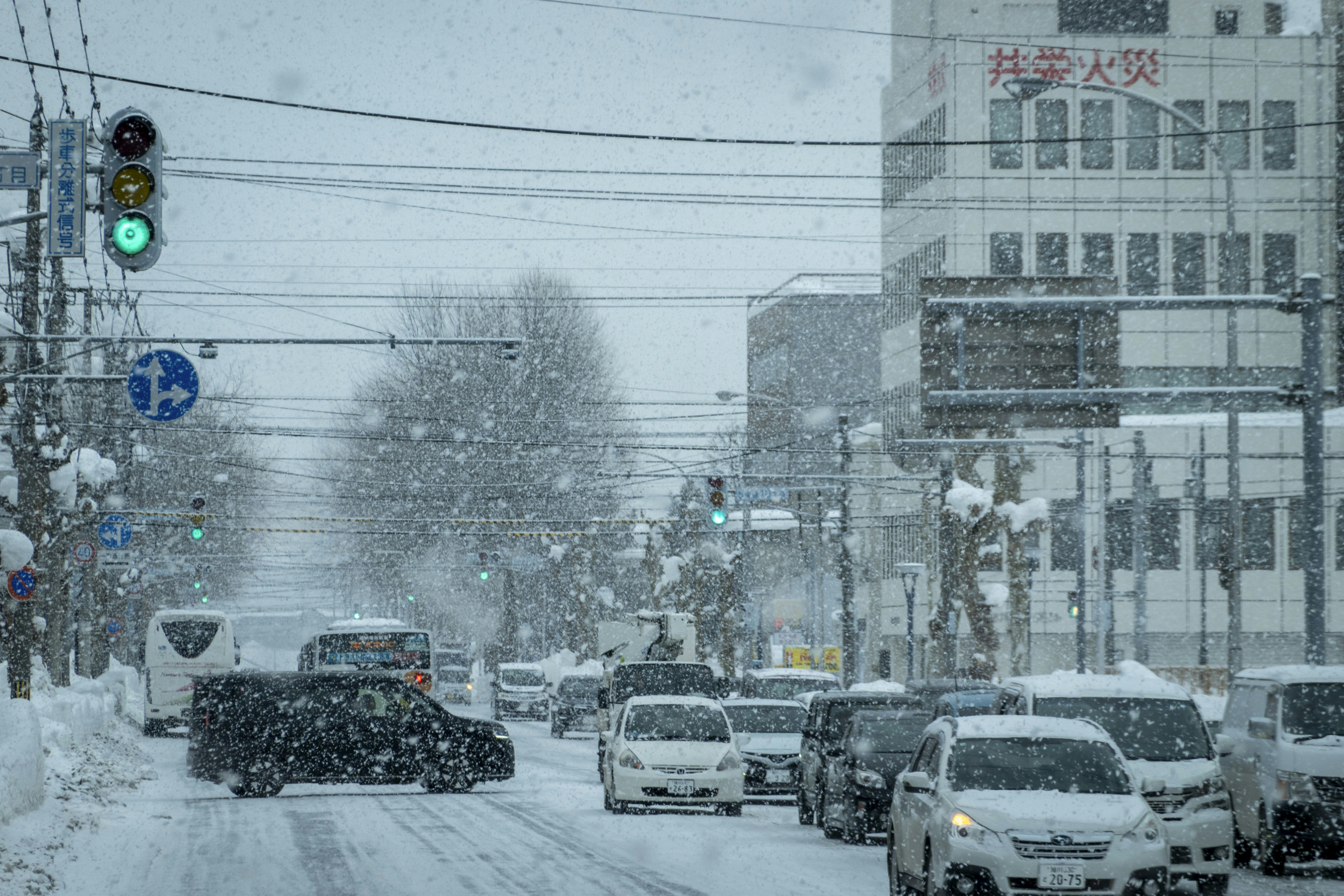 Snow-covered street in Asahikawa with moving traffic and buildings under a cloudy sky.