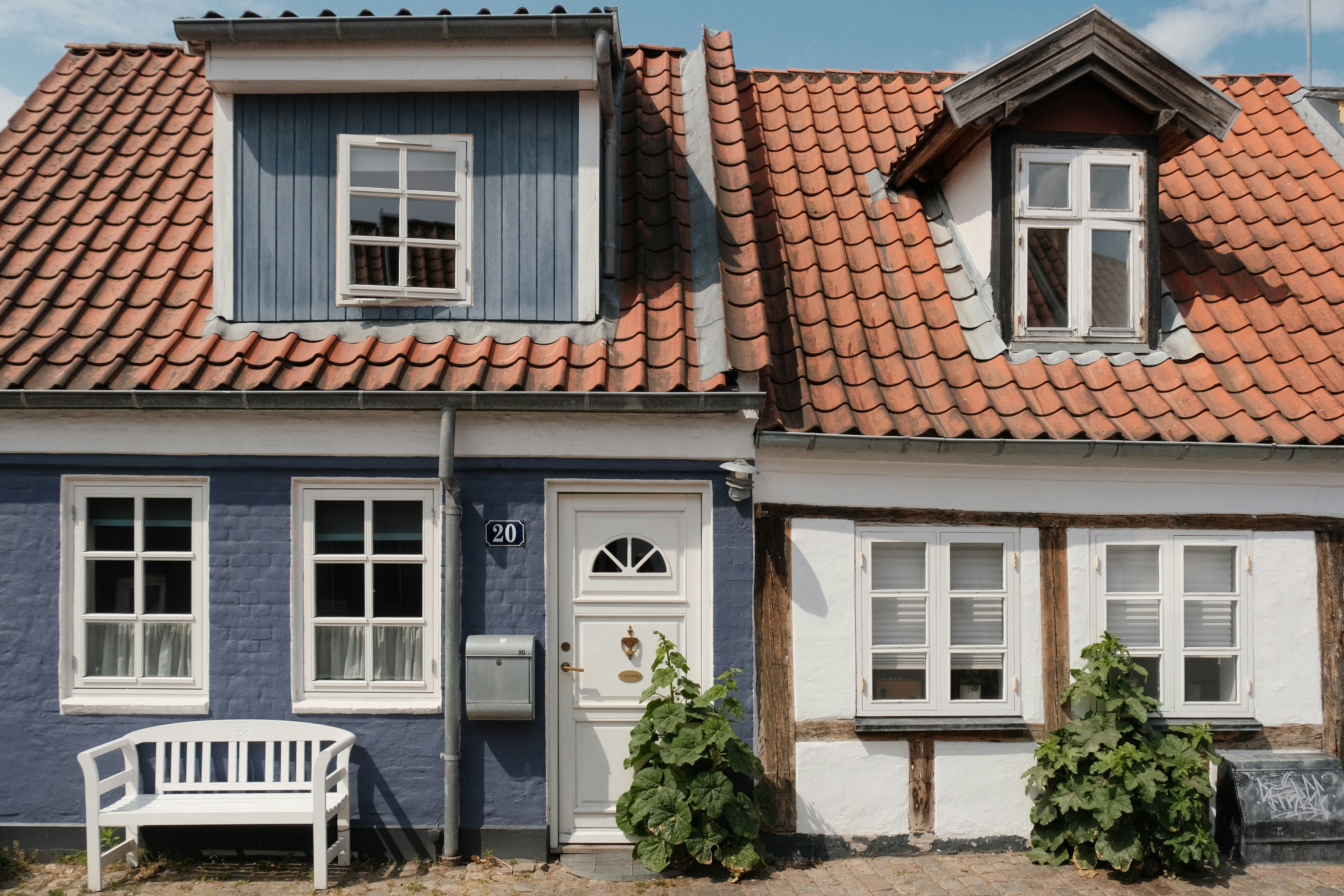 Blue house with red-tiled roof and a white bench in front, flanked by green plants.