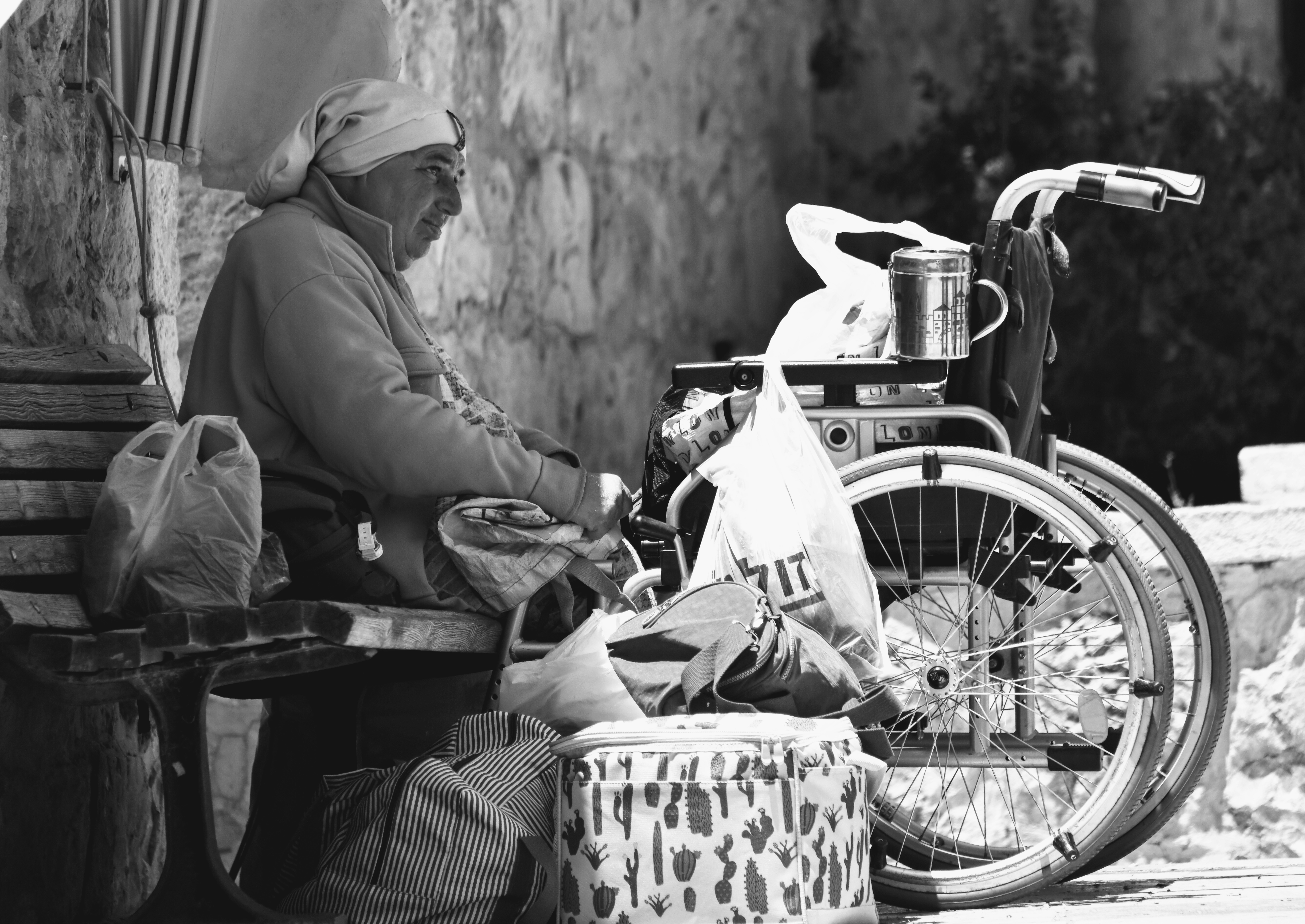 A contemplative figure seated beside a wheelchair, surrounded by bags and personal belongings, reflecting a moment of pause and introspection.