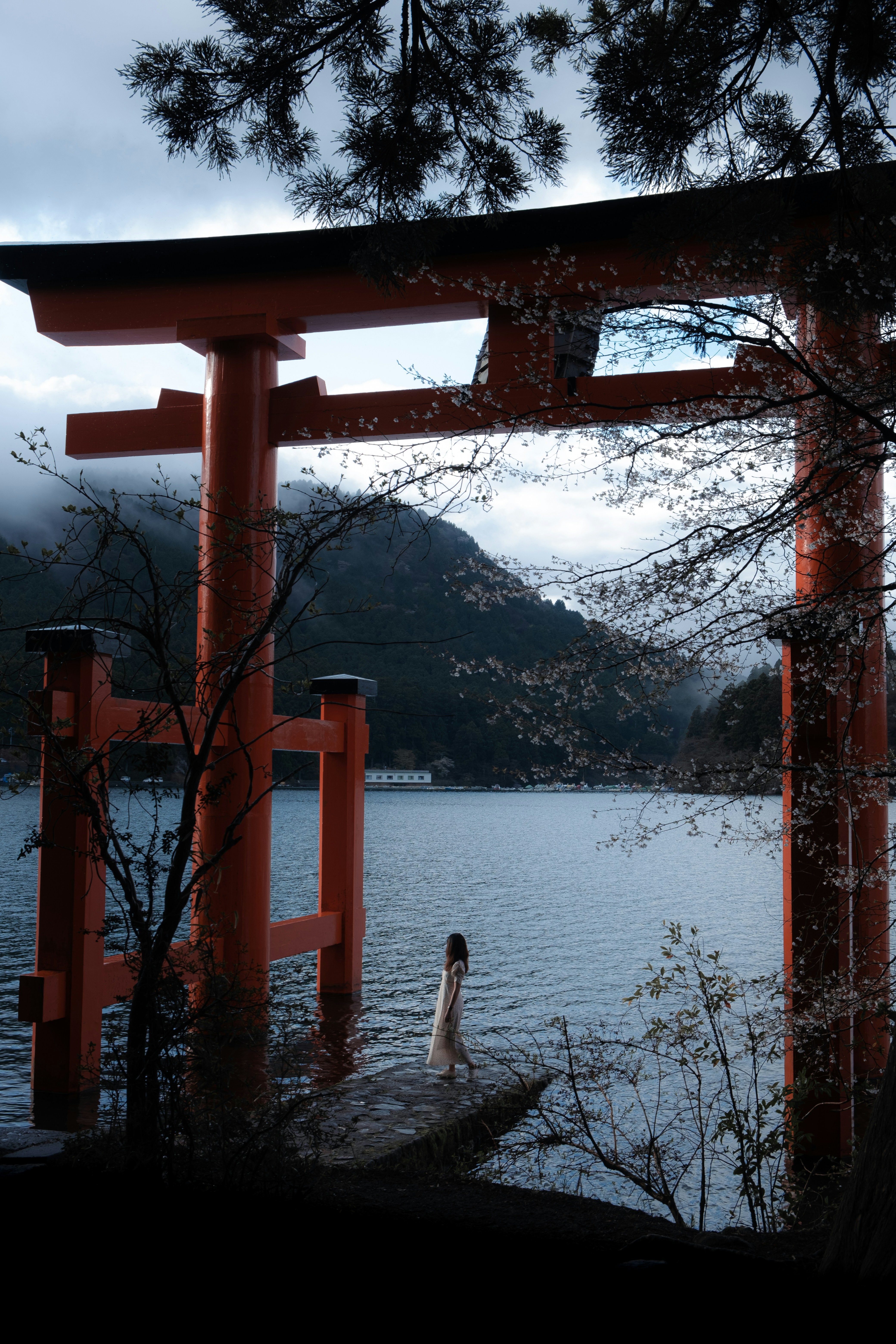 A serene scene featuring a woman walking along the shore near a vibrant torii gate, with misty mountains in the background reflecting on the water's surface.