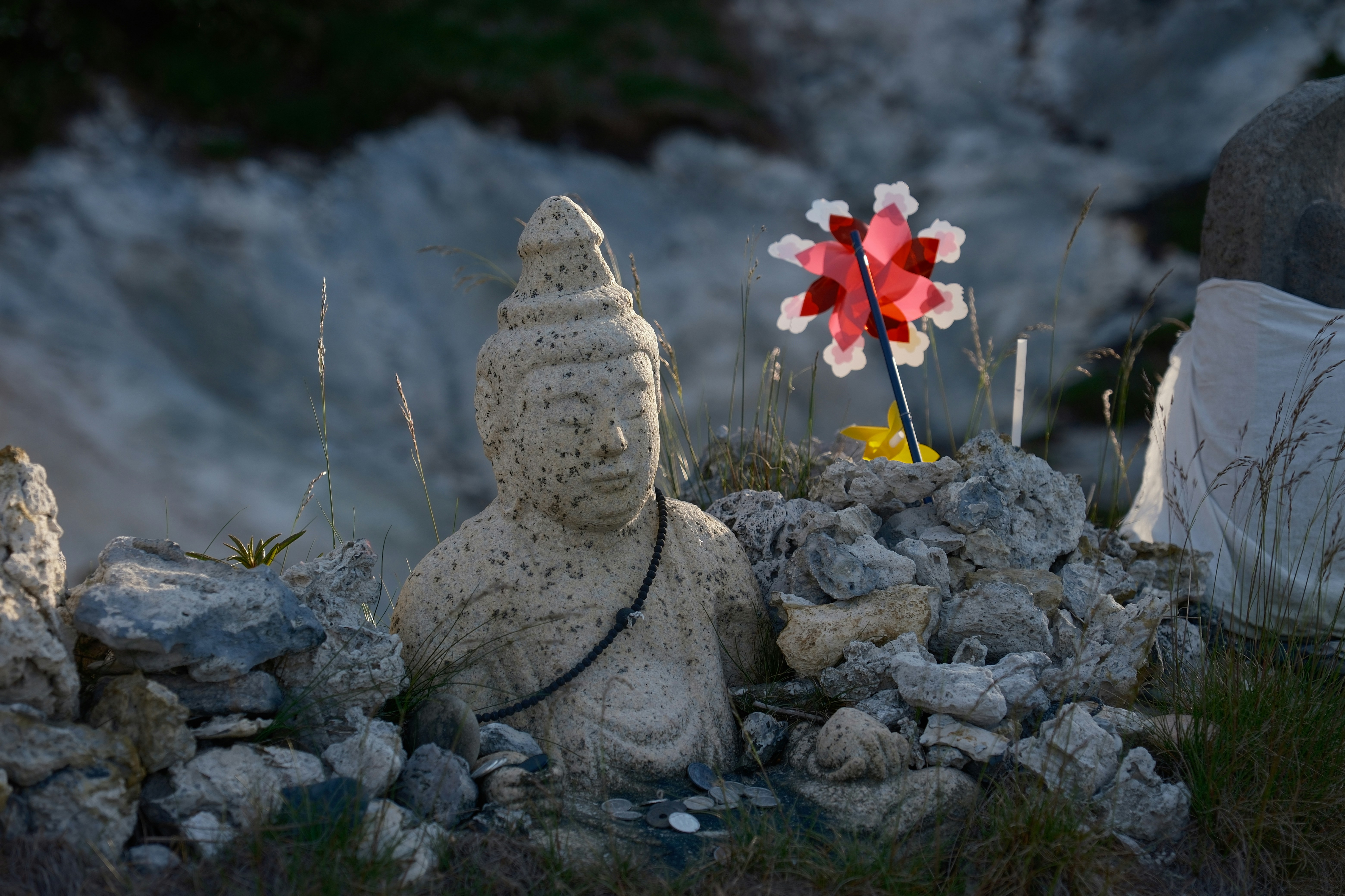 Weathered Buddha Guarding a Pebble Altar with a Red Pinwheel by Quiet Water