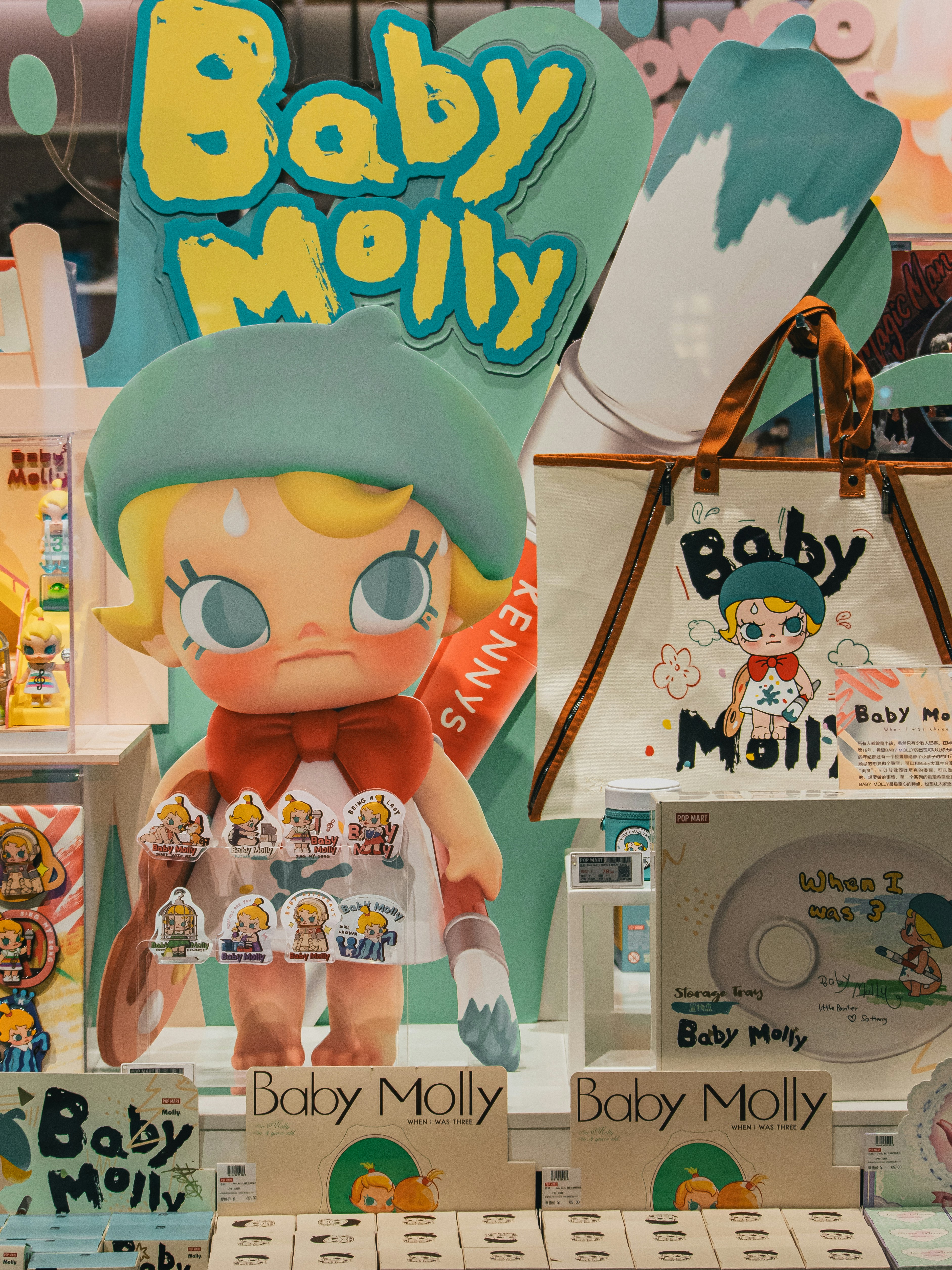 A large Baby Molly figurine anchors a bright toy-store display, with a branded tote showing the Baby Molly logo beside boxed merchandise. The setup highlights character branding and collectible merchandise in retail.