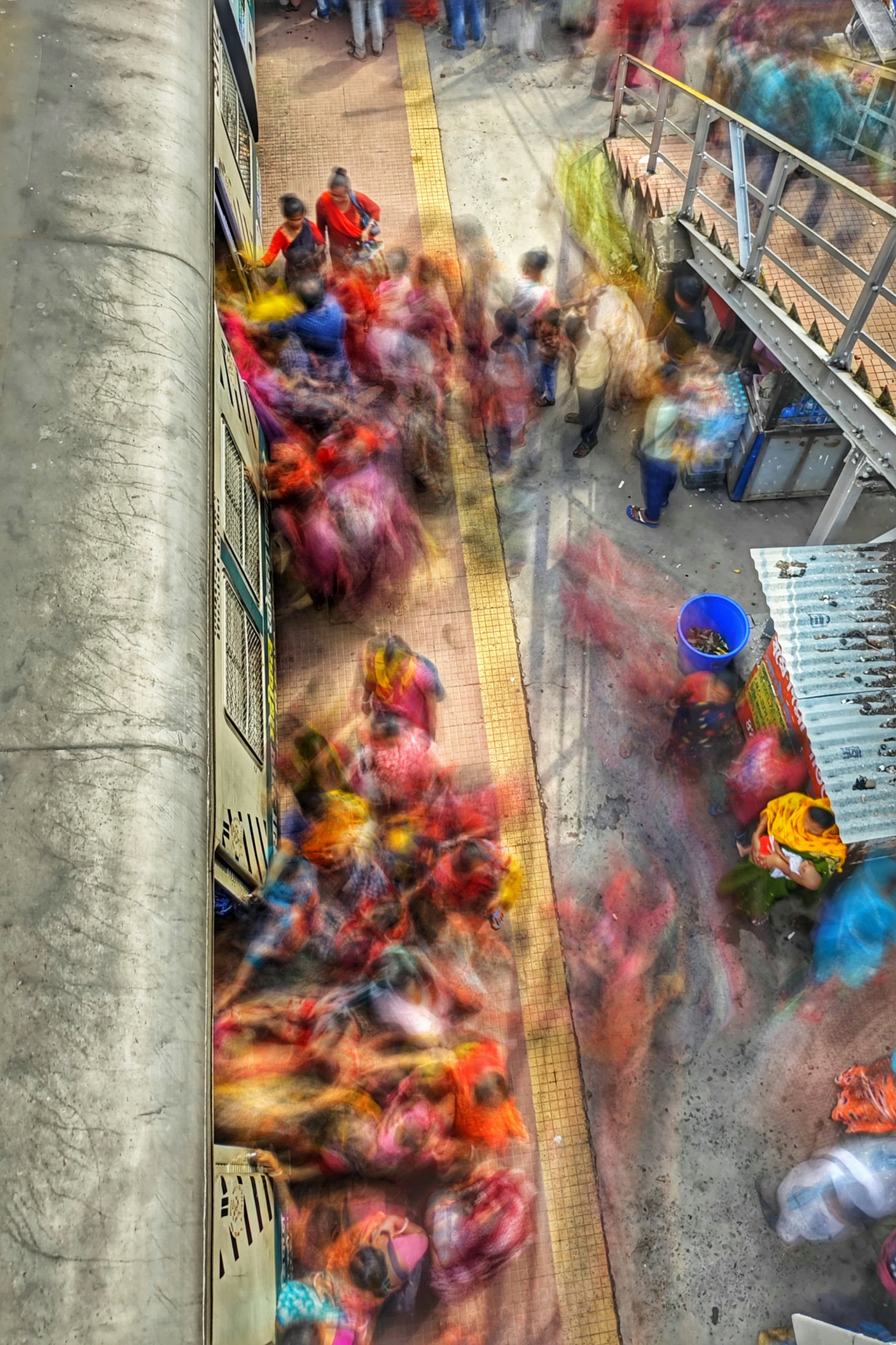 Overhead view of a crowded railway platform where pedestrians blur into color streaks around a central yellow tactile strip.