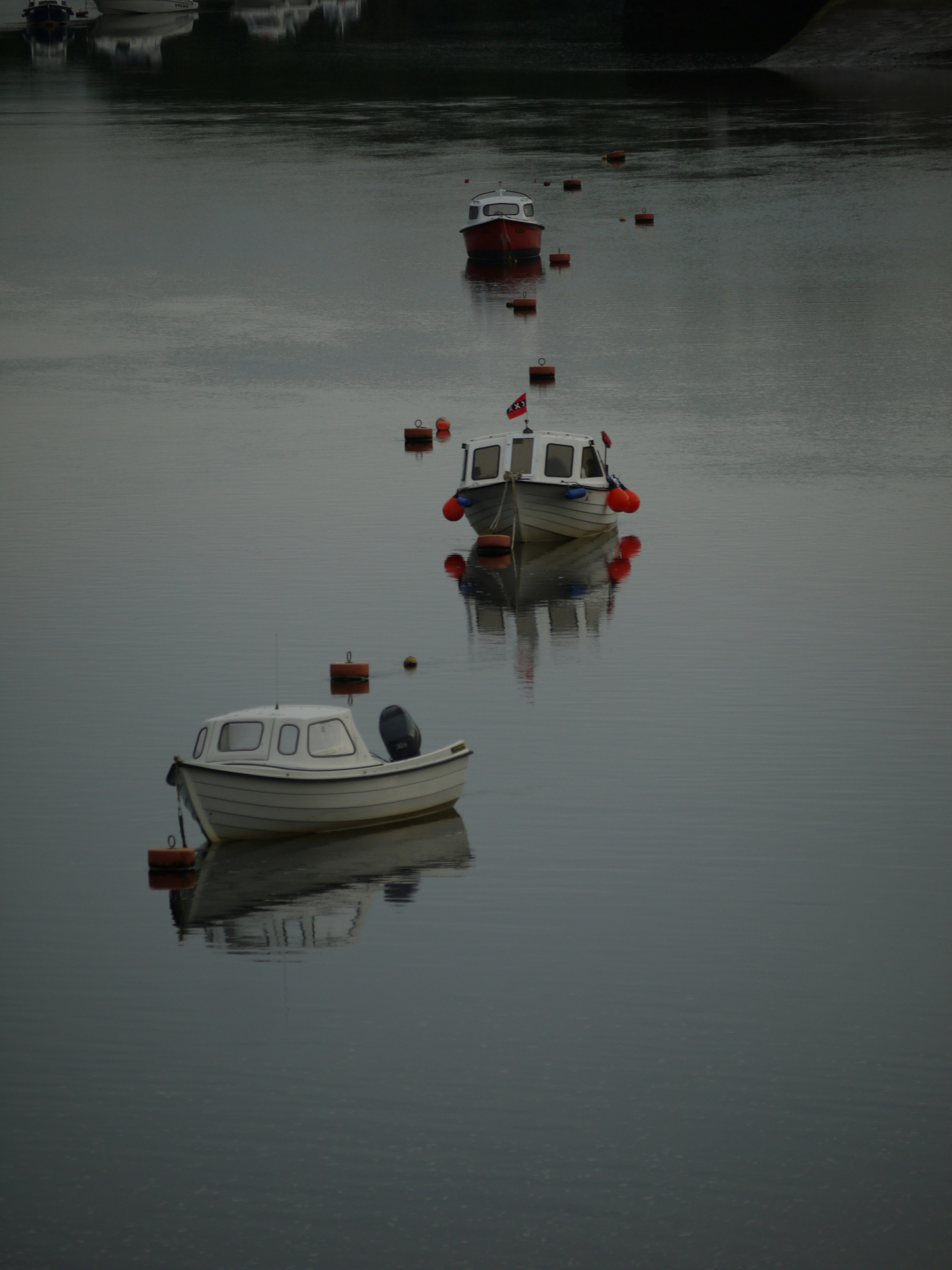 Three boats anchored in calm waters, reflecting their surroundings as dusk settles in. The scene captures a tranquil moment in a harbor.