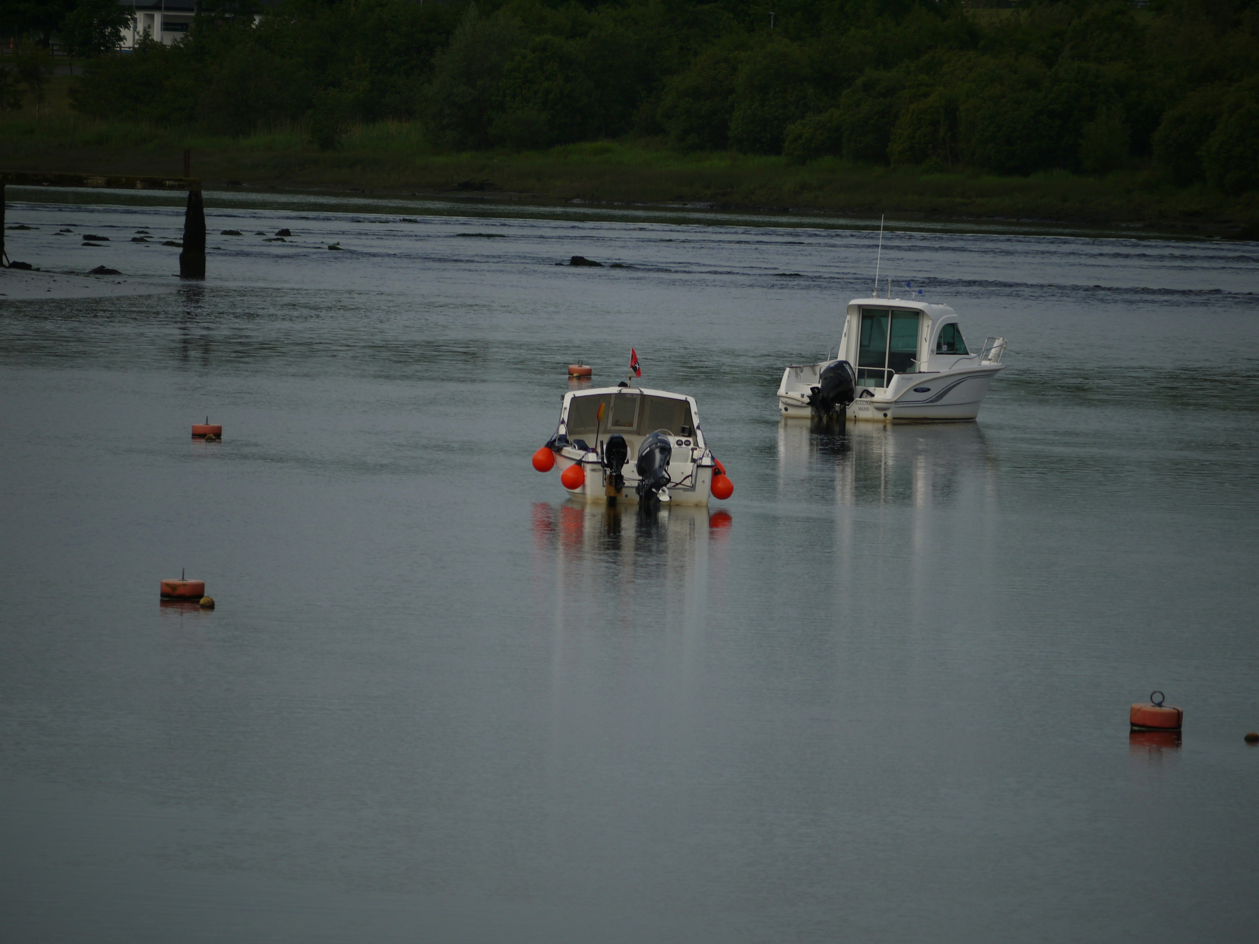 Two small boats anchored in a glassy harbor, with red buoys scattered across the calm water.