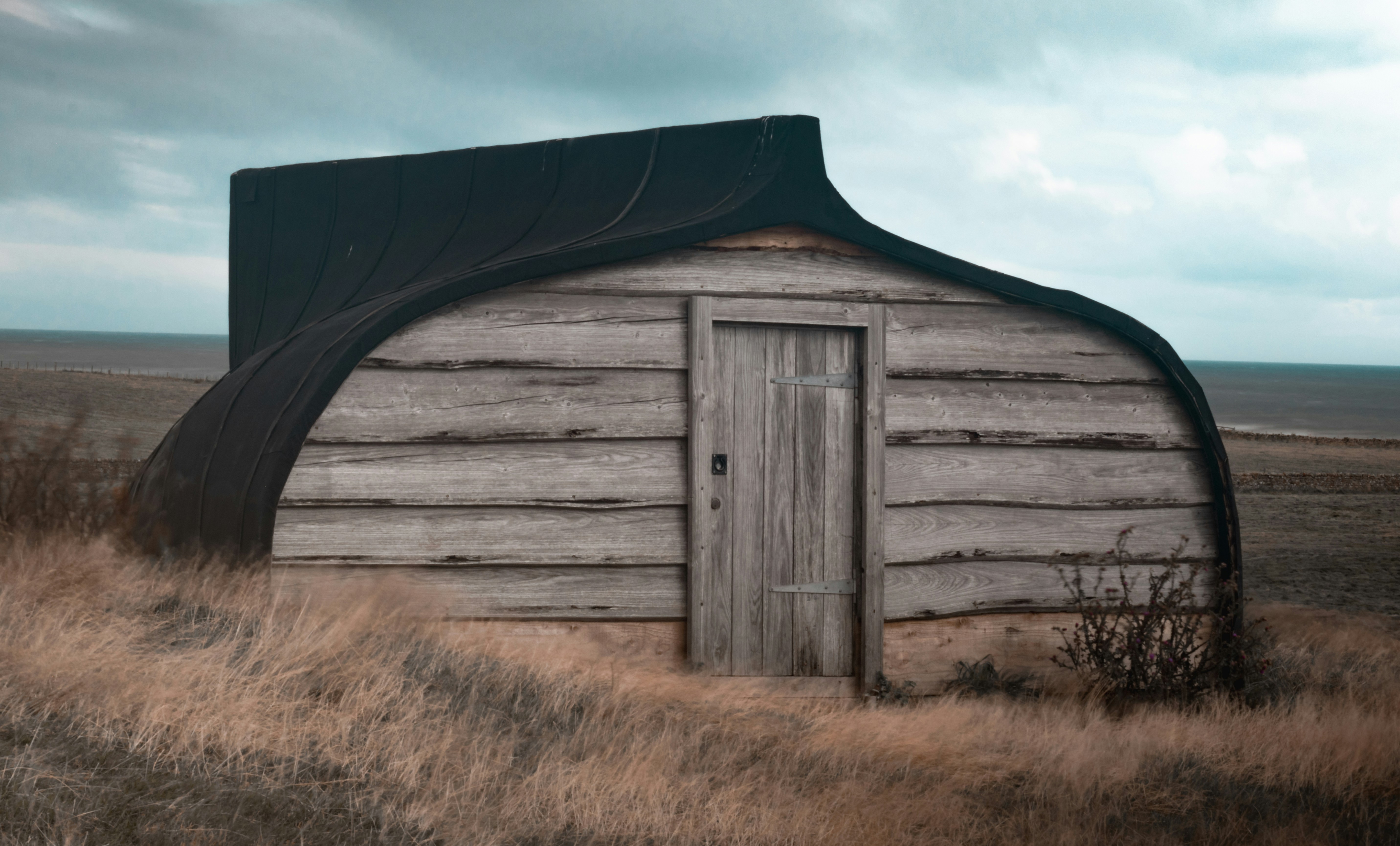 A uniquely shaped wooden cabin stands amidst tall grasses on a vast plain, showcasing its unconventional design against a moody sky.