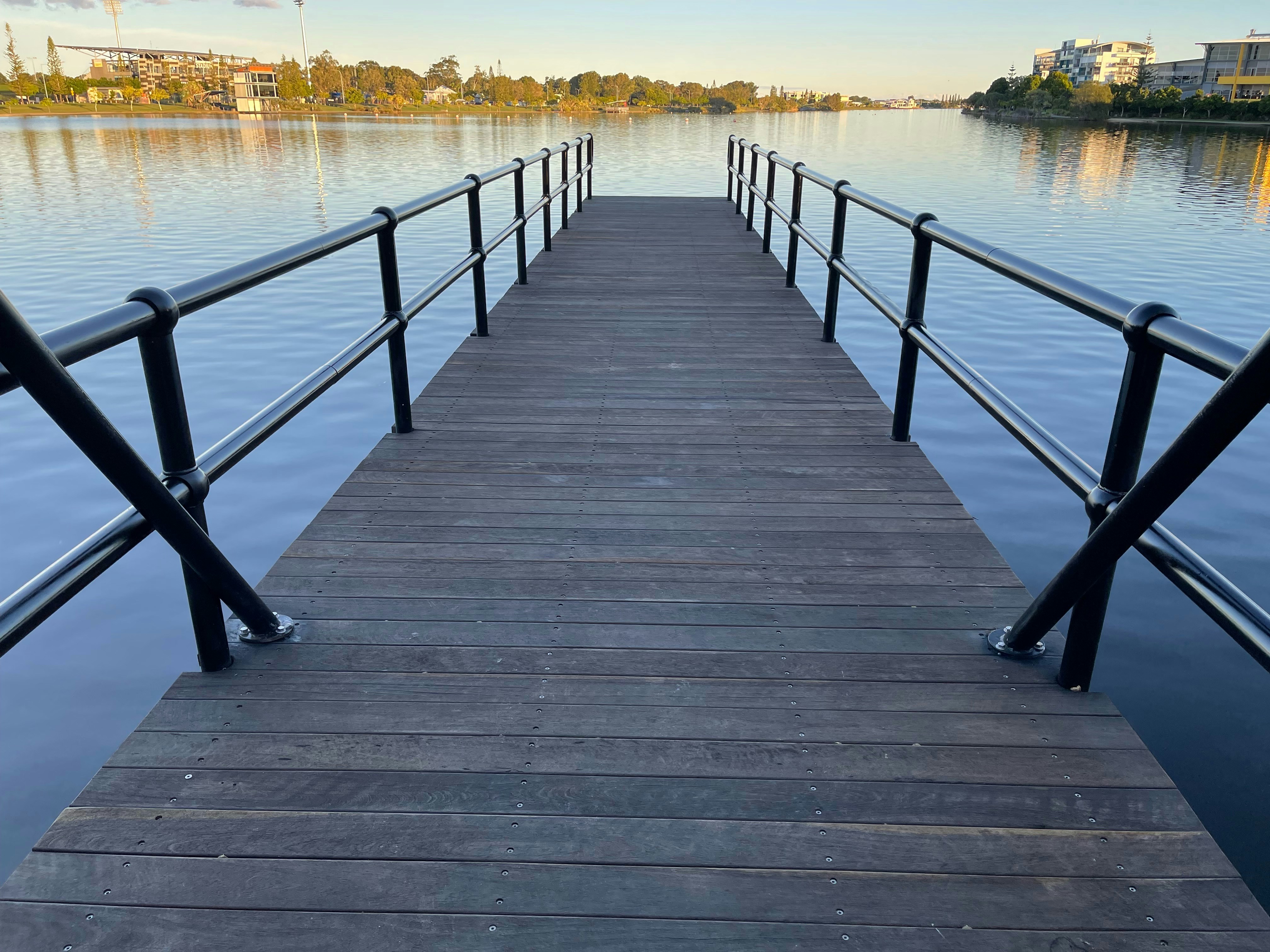 Wooden pier extending over a calm body of water, framed by trees and buildings in the distance.