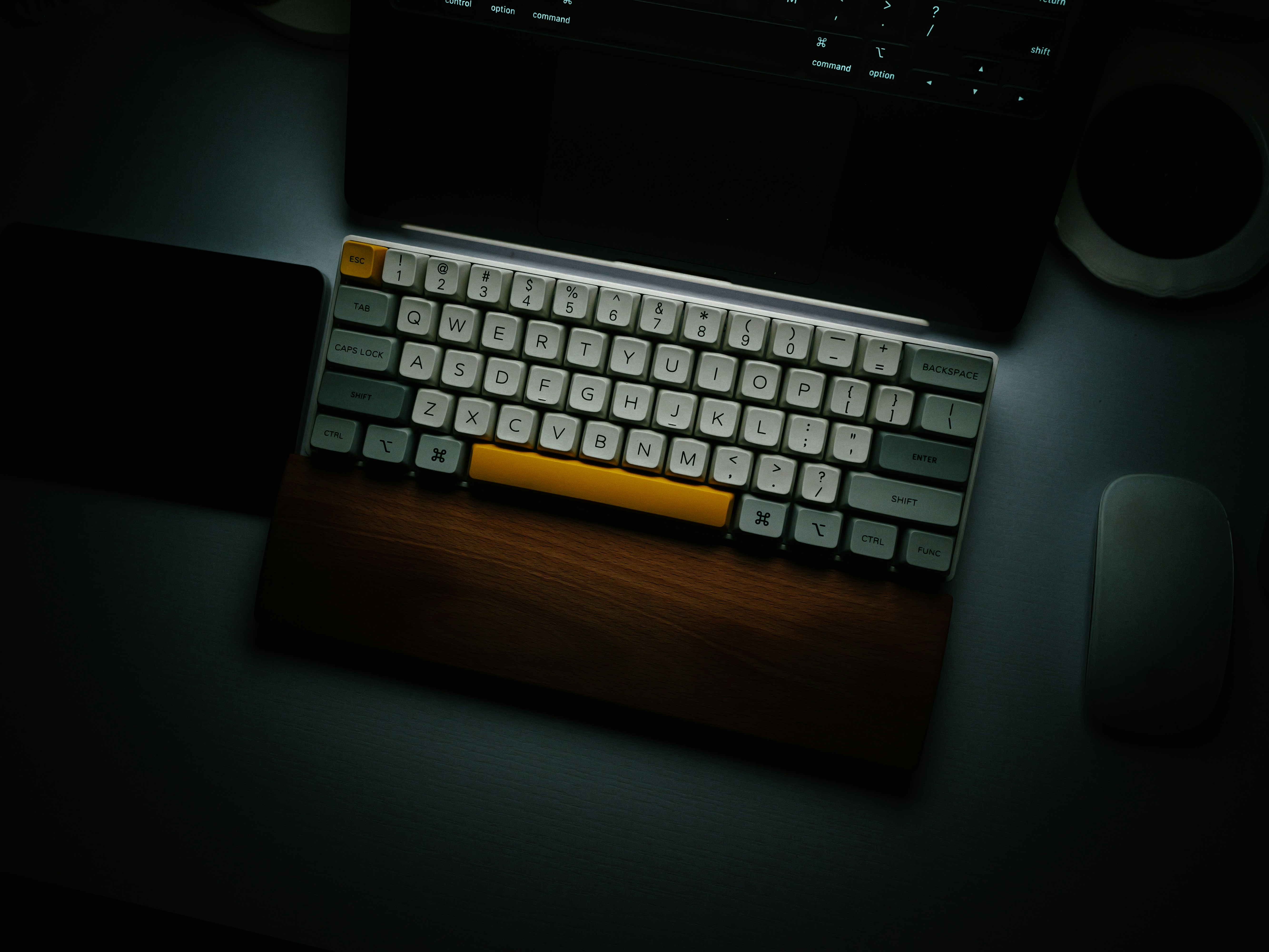 Low-key photograph of a white keyboard with a bright yellow accent bar resting on a wooden palm rest, illuminated by the dim glow of a nearby monitor.