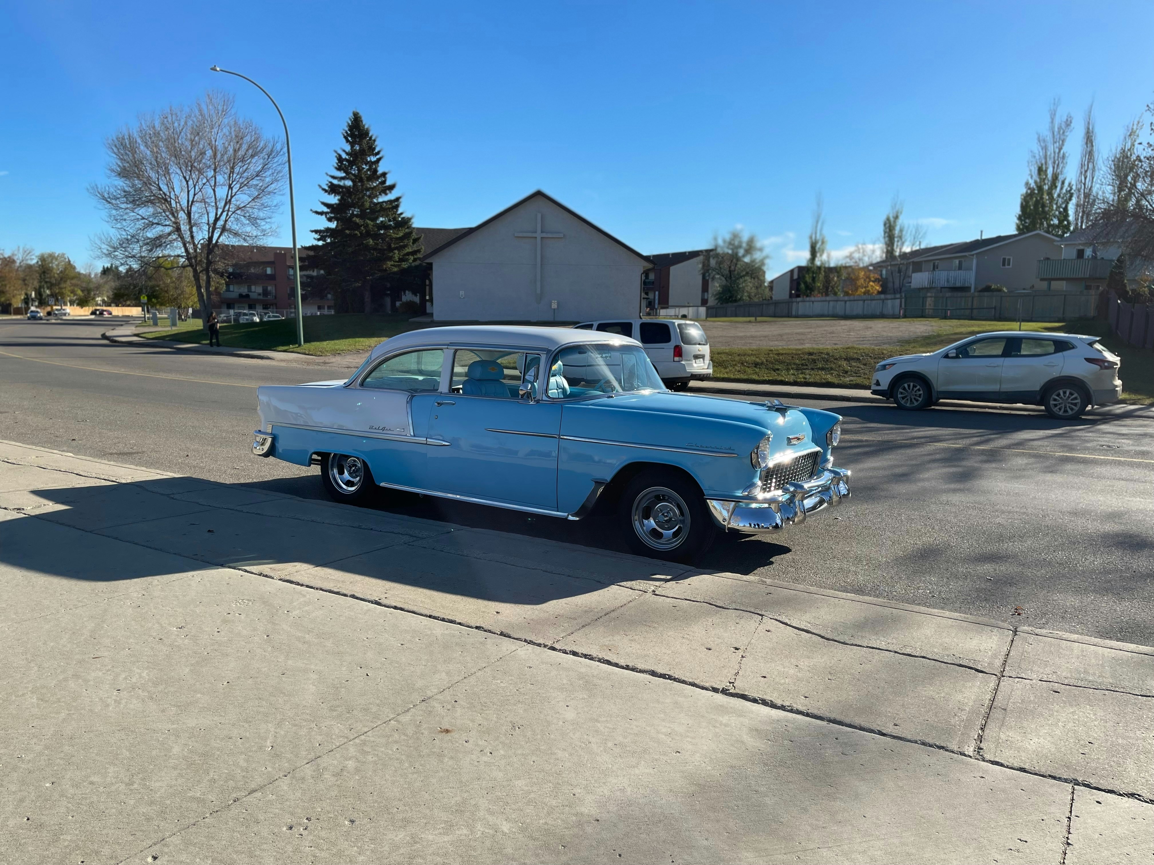 A classic light blue 1955 Chevrolet parked on a suburban road under a clear blue sky.