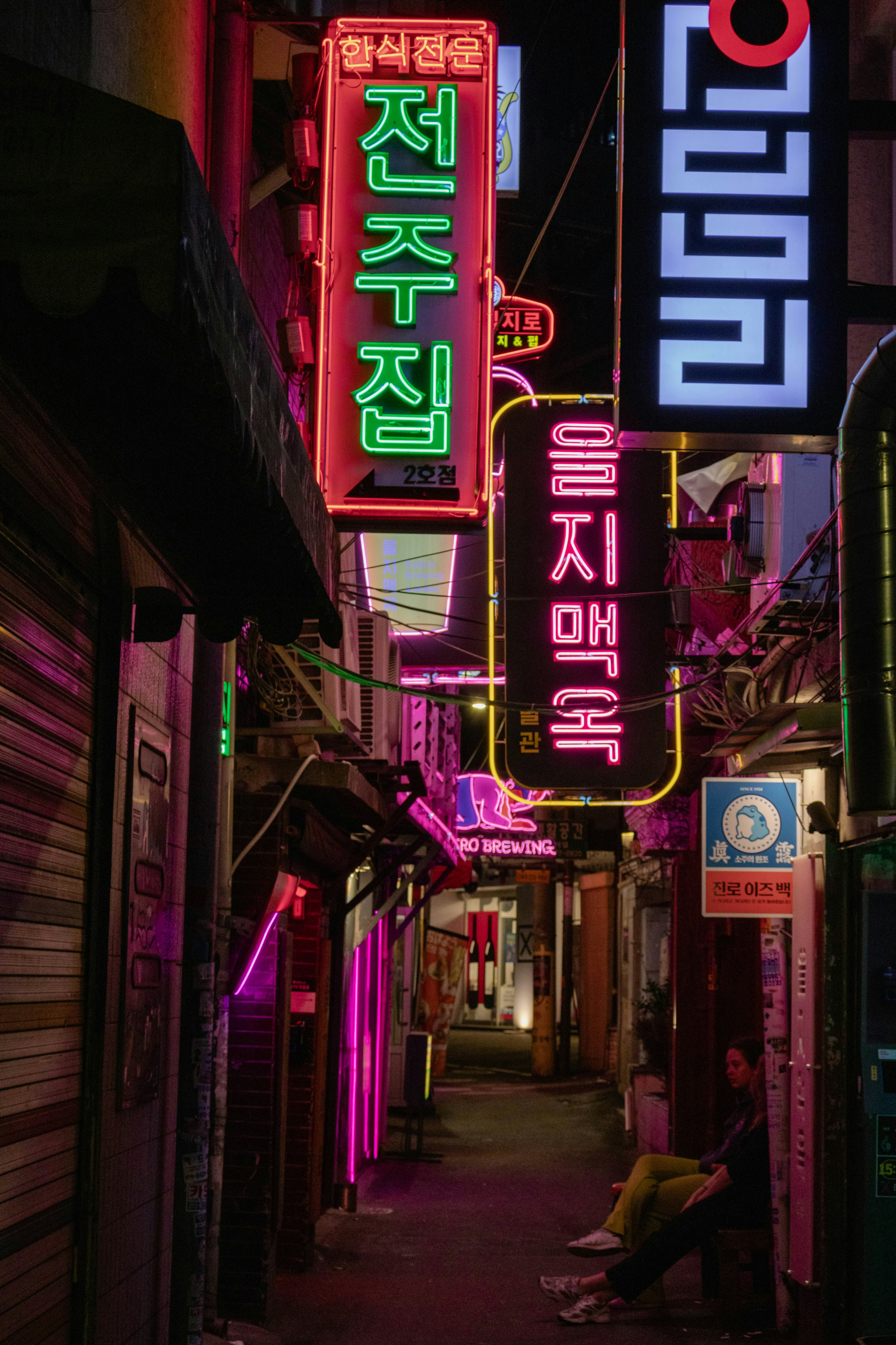 A narrow alley with neon signs and a person sitting on a bench photo ...
