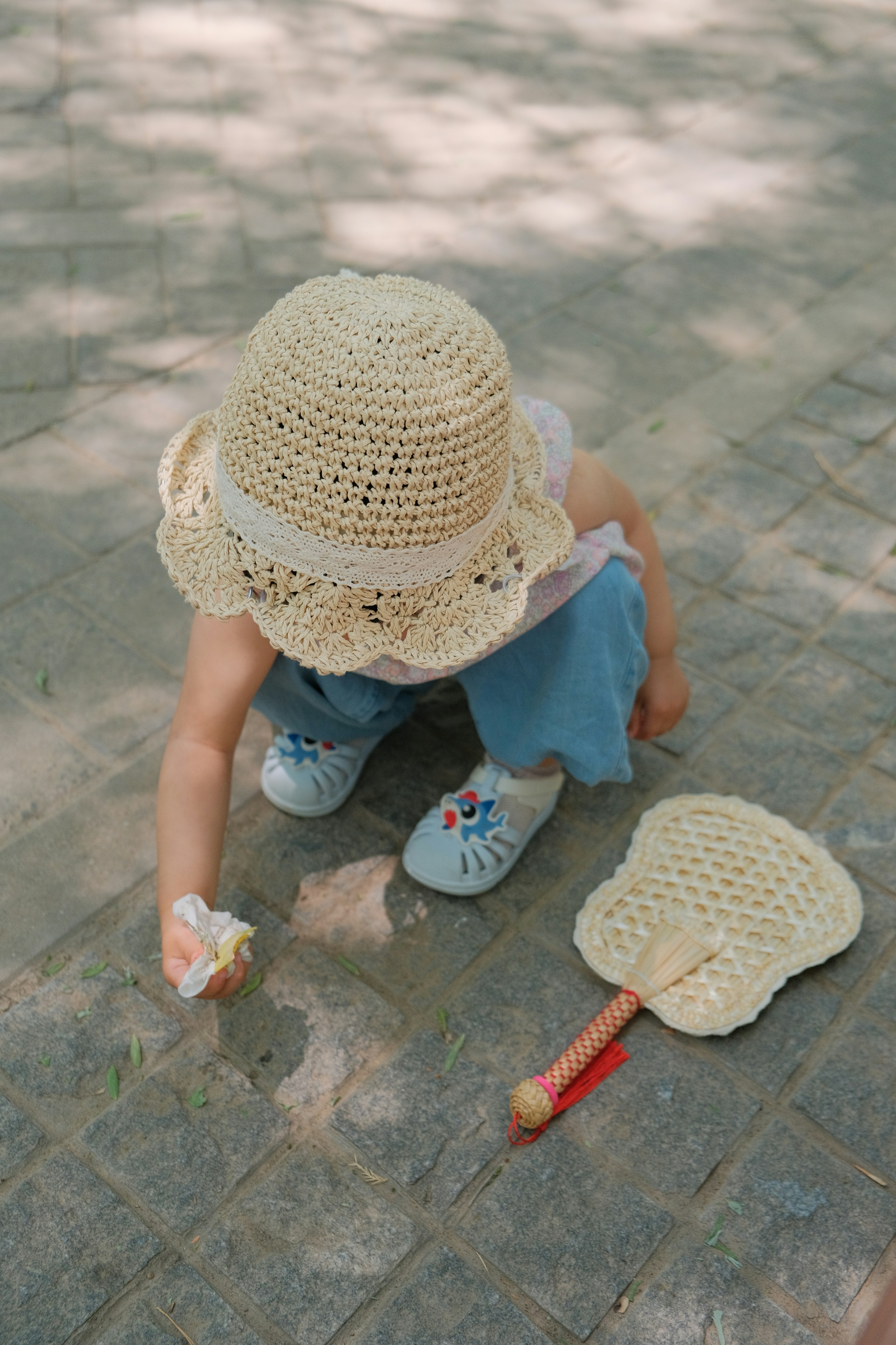 Sunlit Courtyard Discovery: A Little One in a Straw Hat Examines a Tiny Find