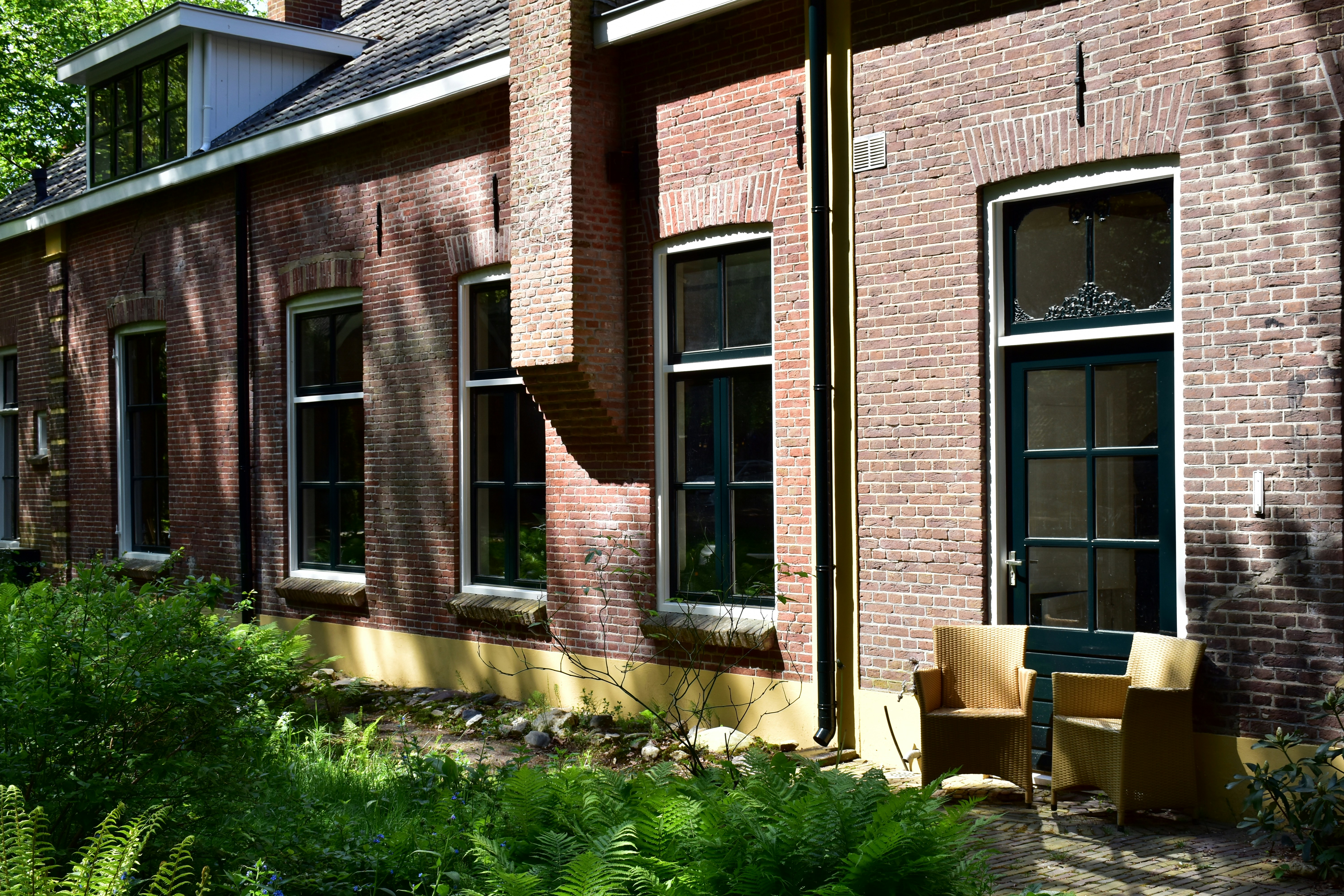 Sunlit red brick house exterior with dark green framed windows and garden foliage in the foreground.
