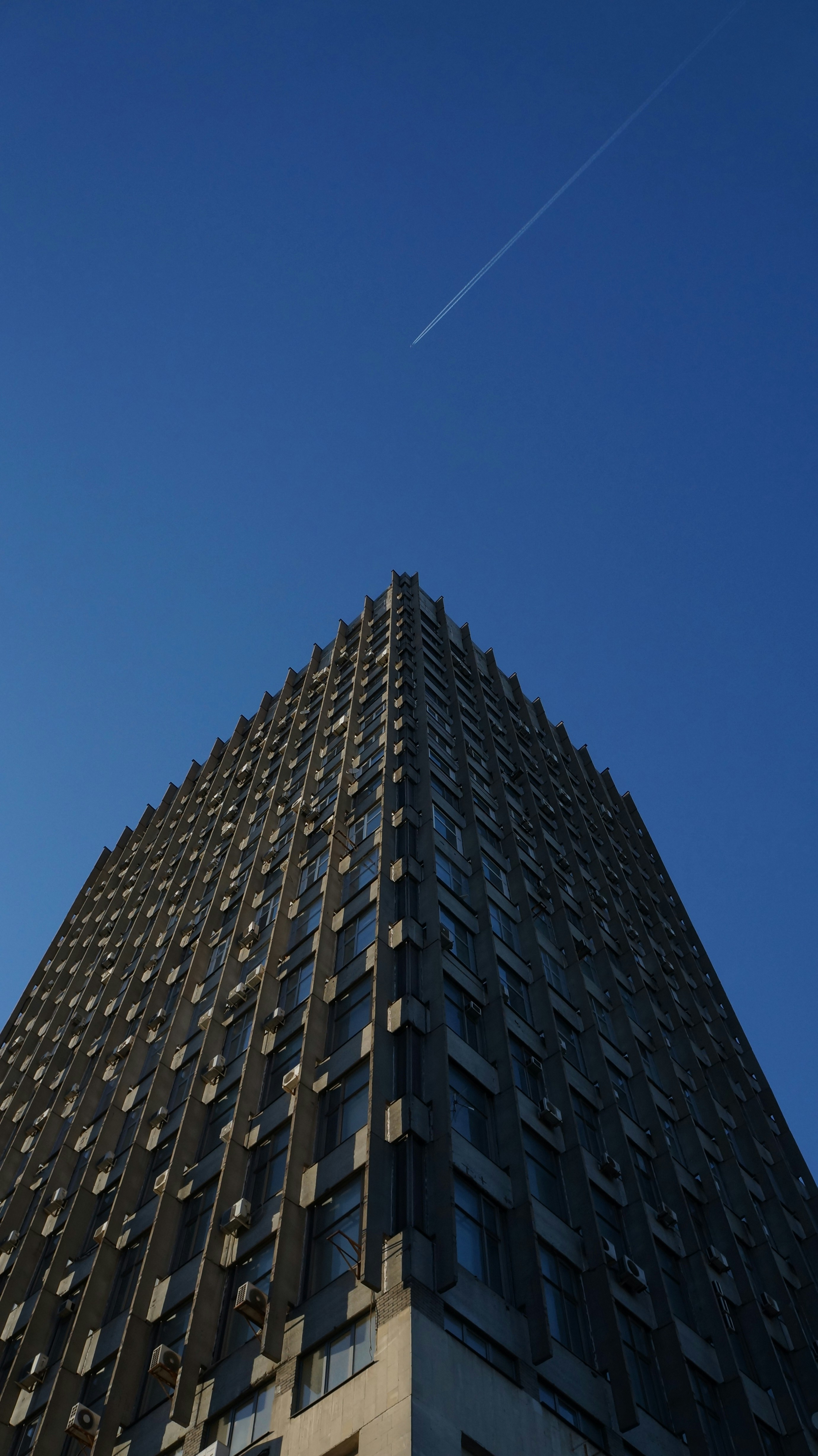 Photograph of a tall office tower with a grid of windows rising against a deep blue sky, a contrail diagonally overhead.