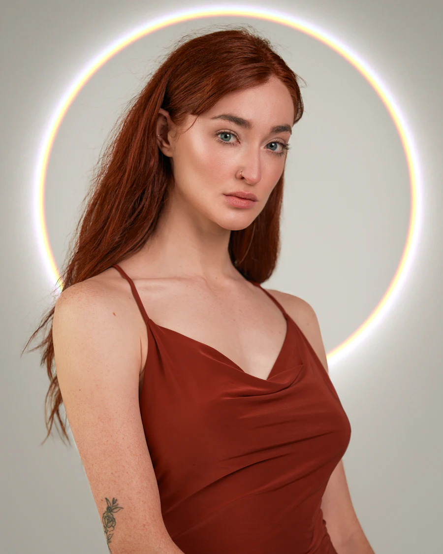 Red-haired woman posing in a rust-colored dress against a pale background, framed by a glowing ring light halo. She looks directly at the camera with a calm expression.