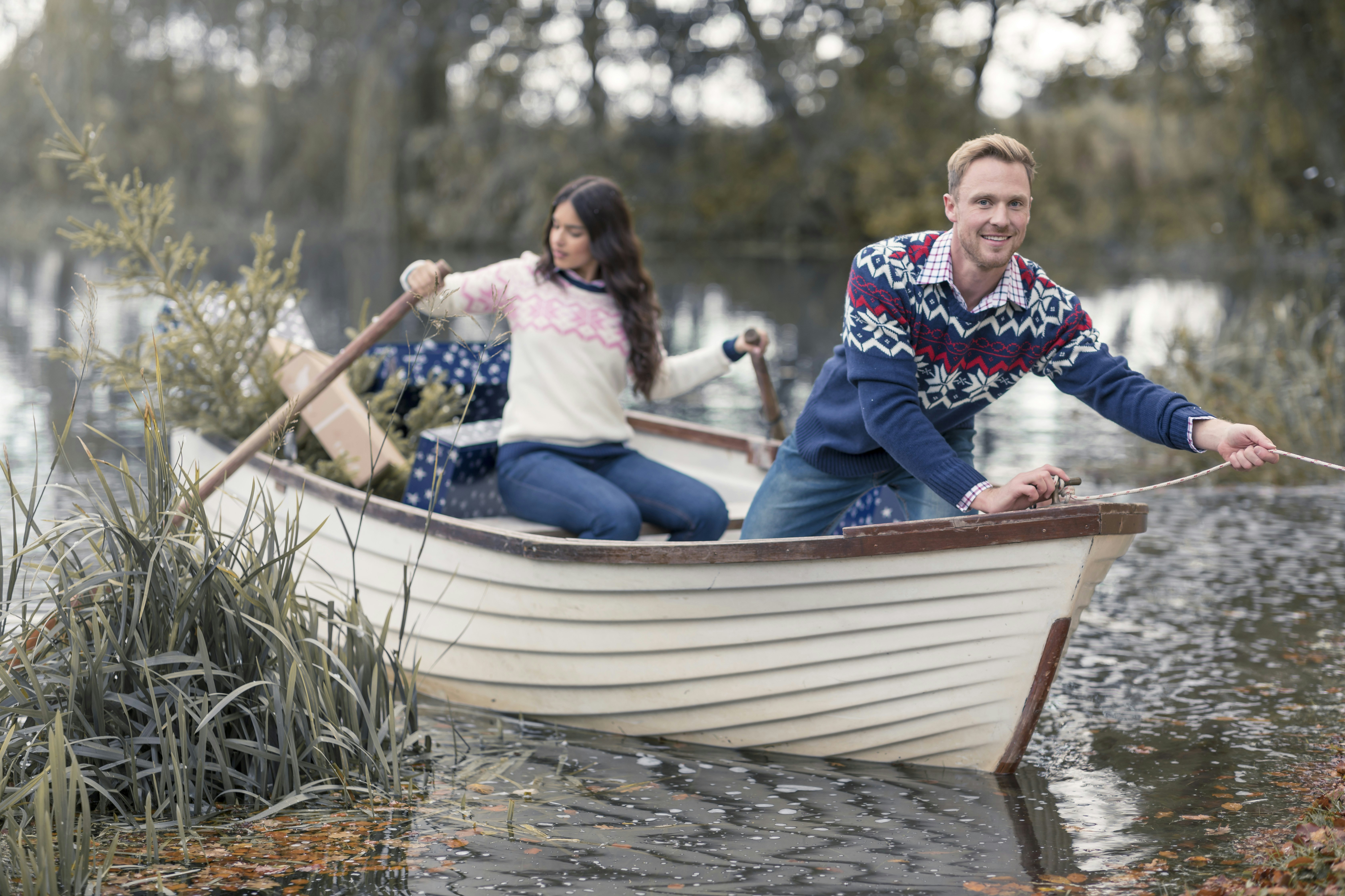 Reed-Lined Drift: Two knit-clad rowers glide a wooden boat across a tranquil lake