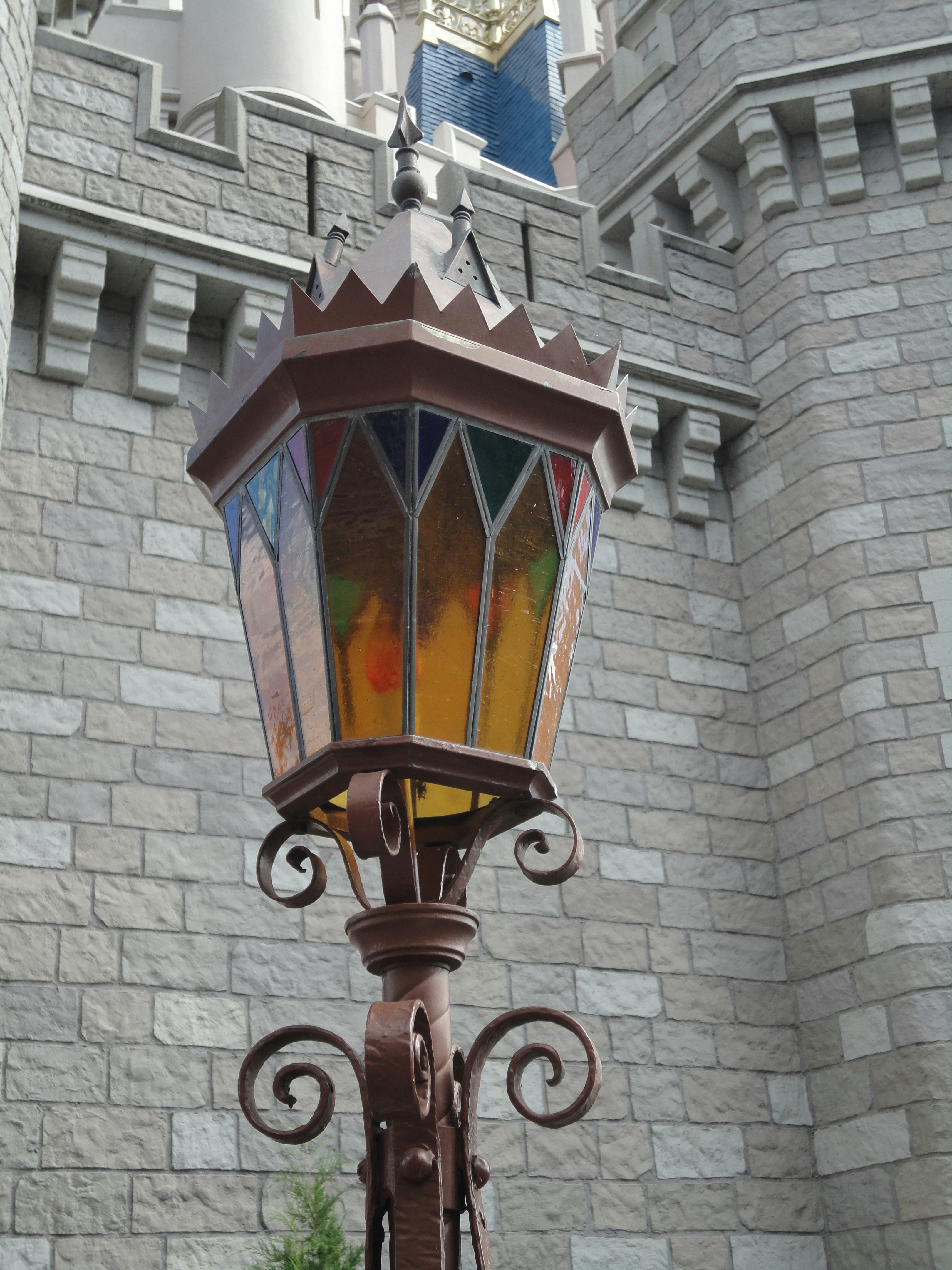 Ornate iron streetlamp with multicolored amber glass panes stands against a textured castle stone wall.