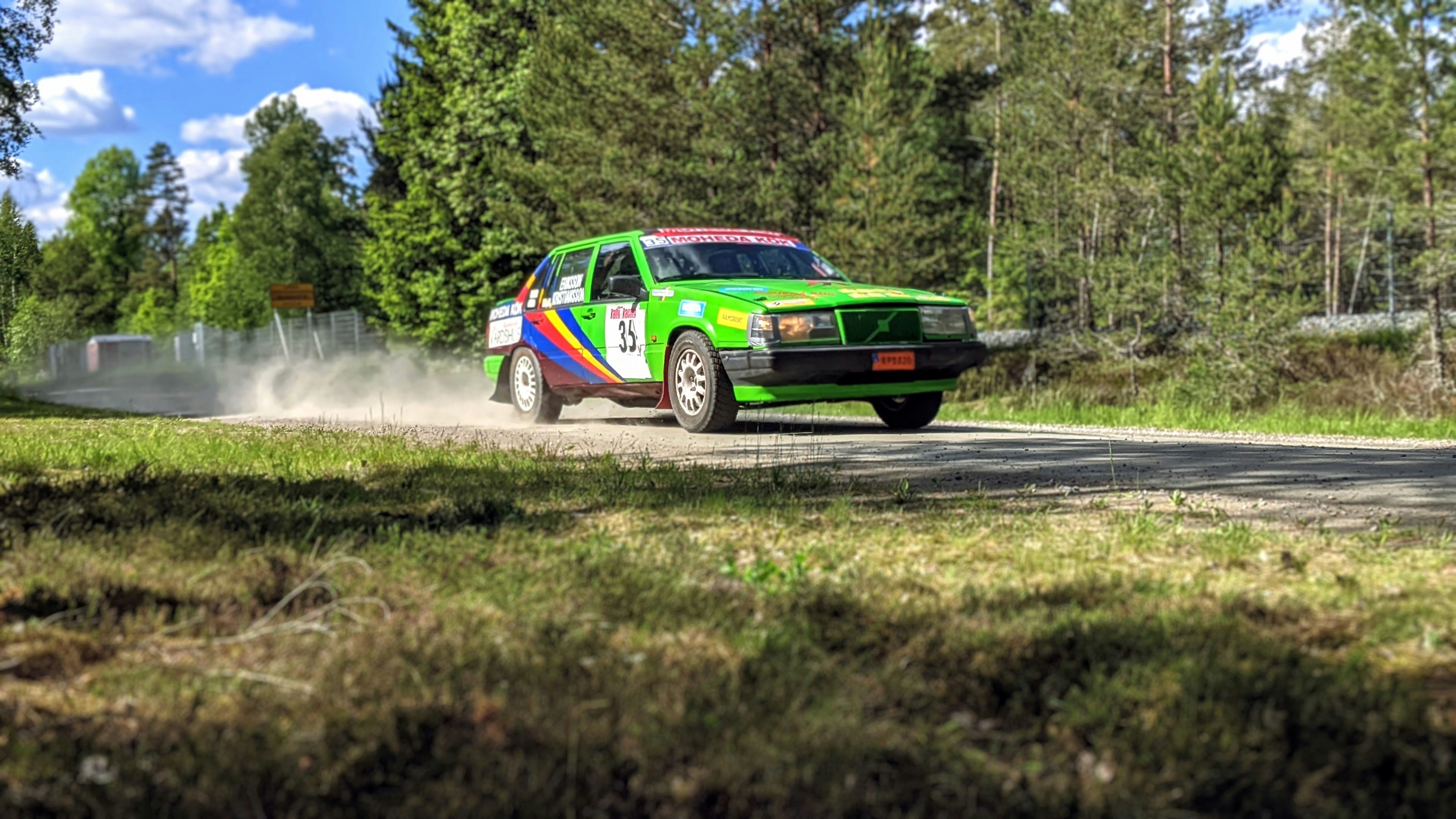A vibrant green rally car speeds down a dirt road, kicking up dust amidst a backdrop of lush trees and blue skies.