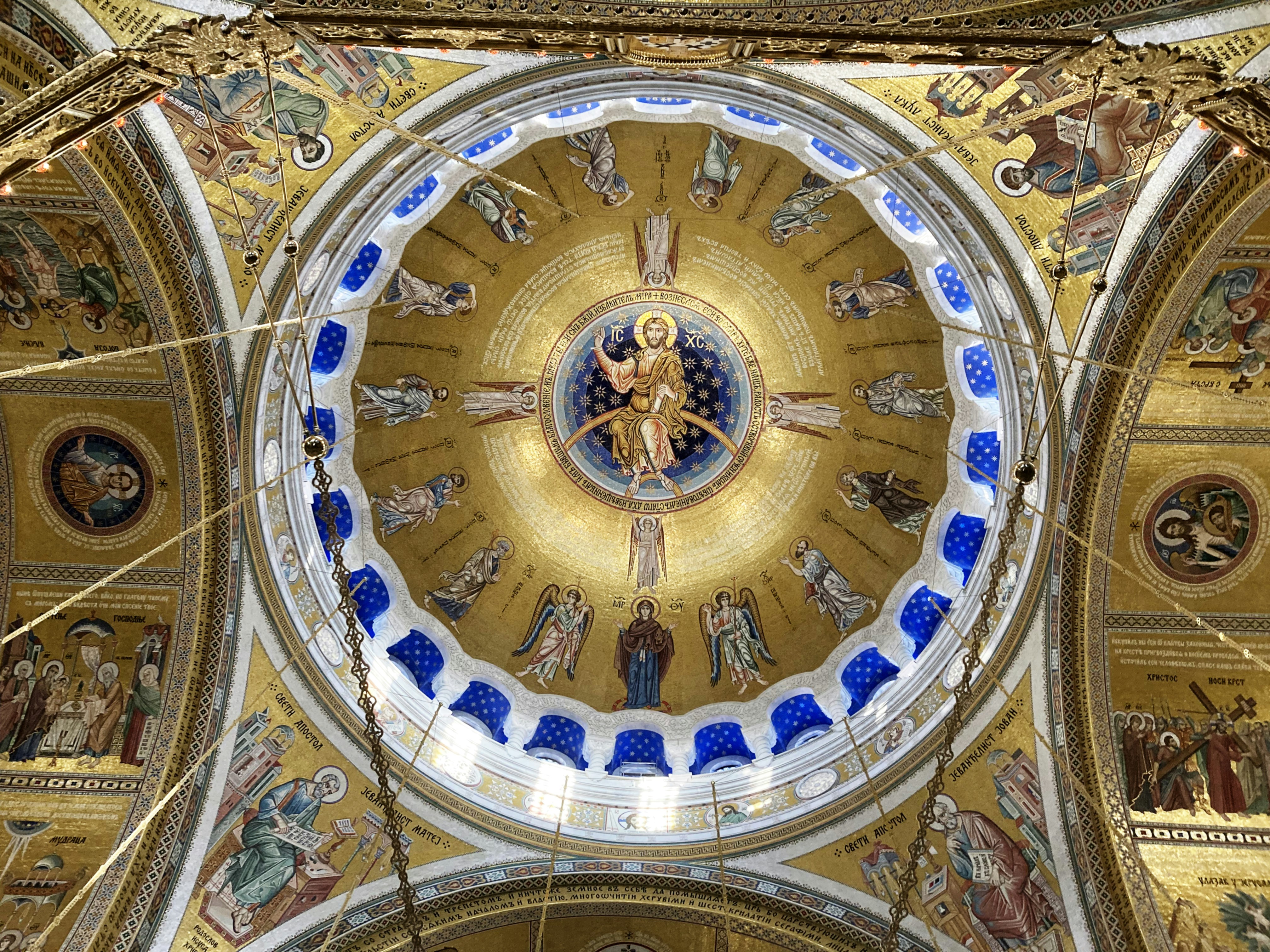 Intricate frescoes and golden mosaics adorn the dome of a historic church, showcasing religious figures and celestial themes.