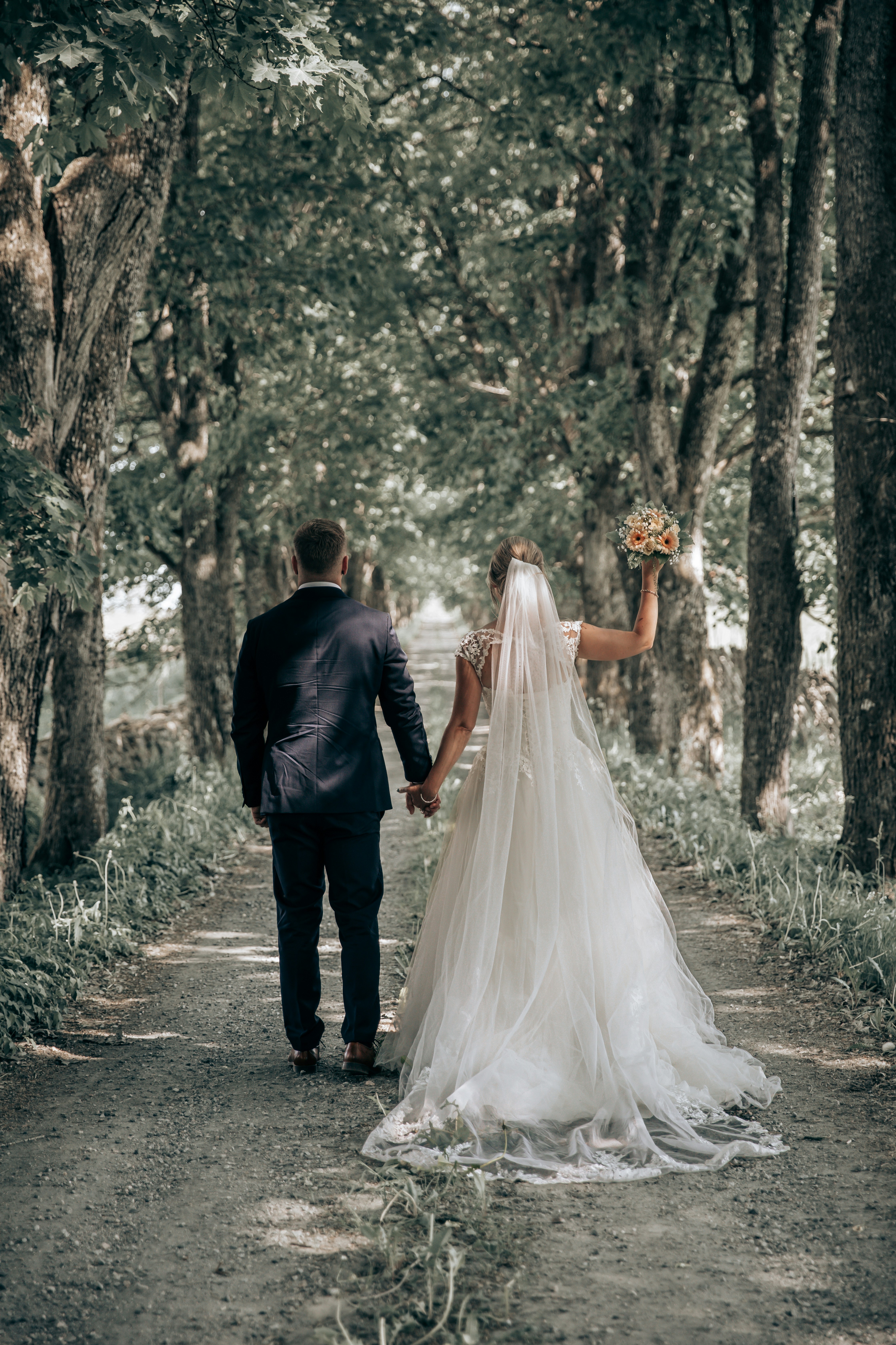 Couple walking hand in hand down a tree-lined path, with the bride holding a bouquet aloft. The serene atmosphere enhances the romantic moment.