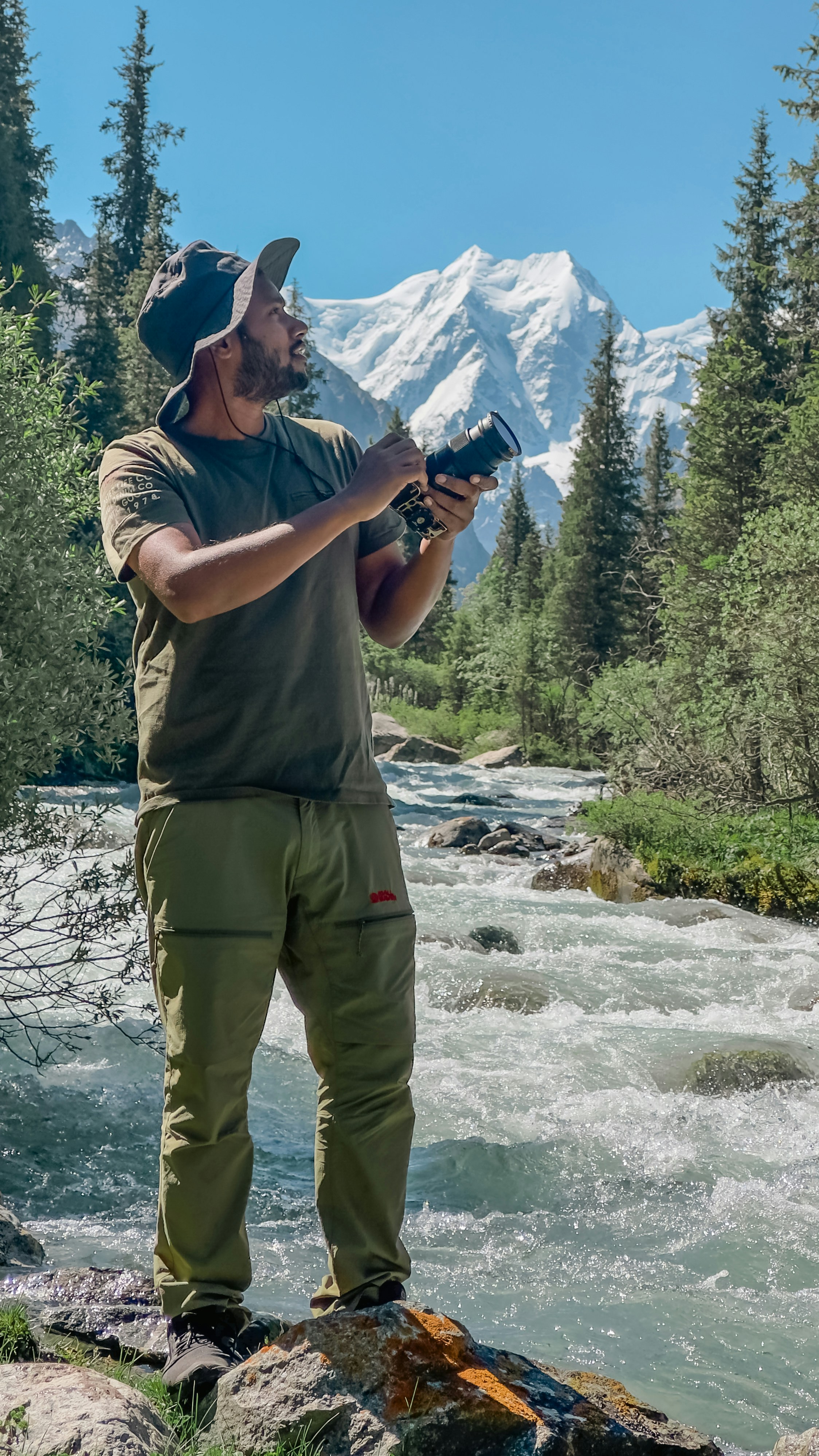A photographer stands in a forest beside a rushing river, mountain peaks in the background, holding a camera and lens ready to shoot. The scene captures outdoor exploration; a logo-free, natural setting.