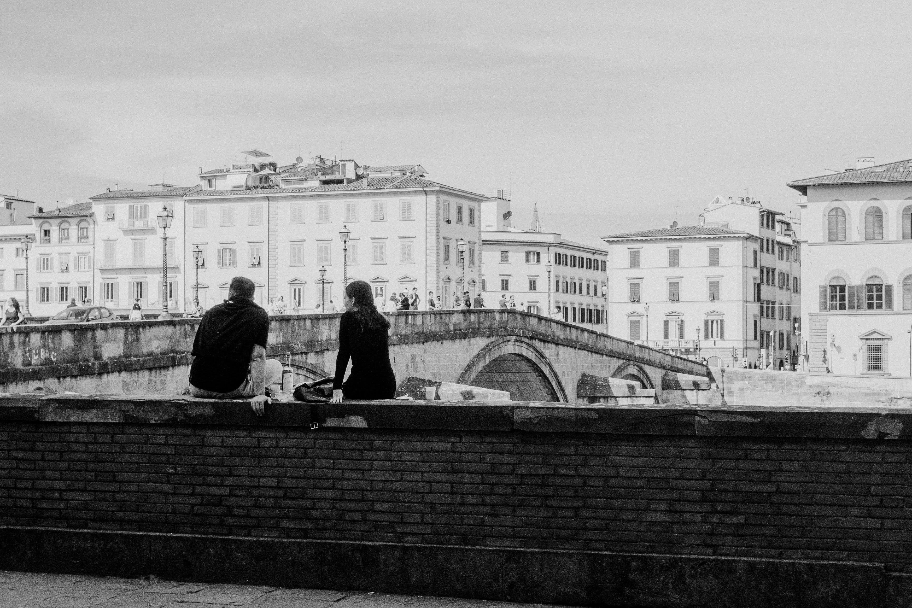 Two people sit on a stone wall overlooking a bridge and historic buildings in a cityscape.