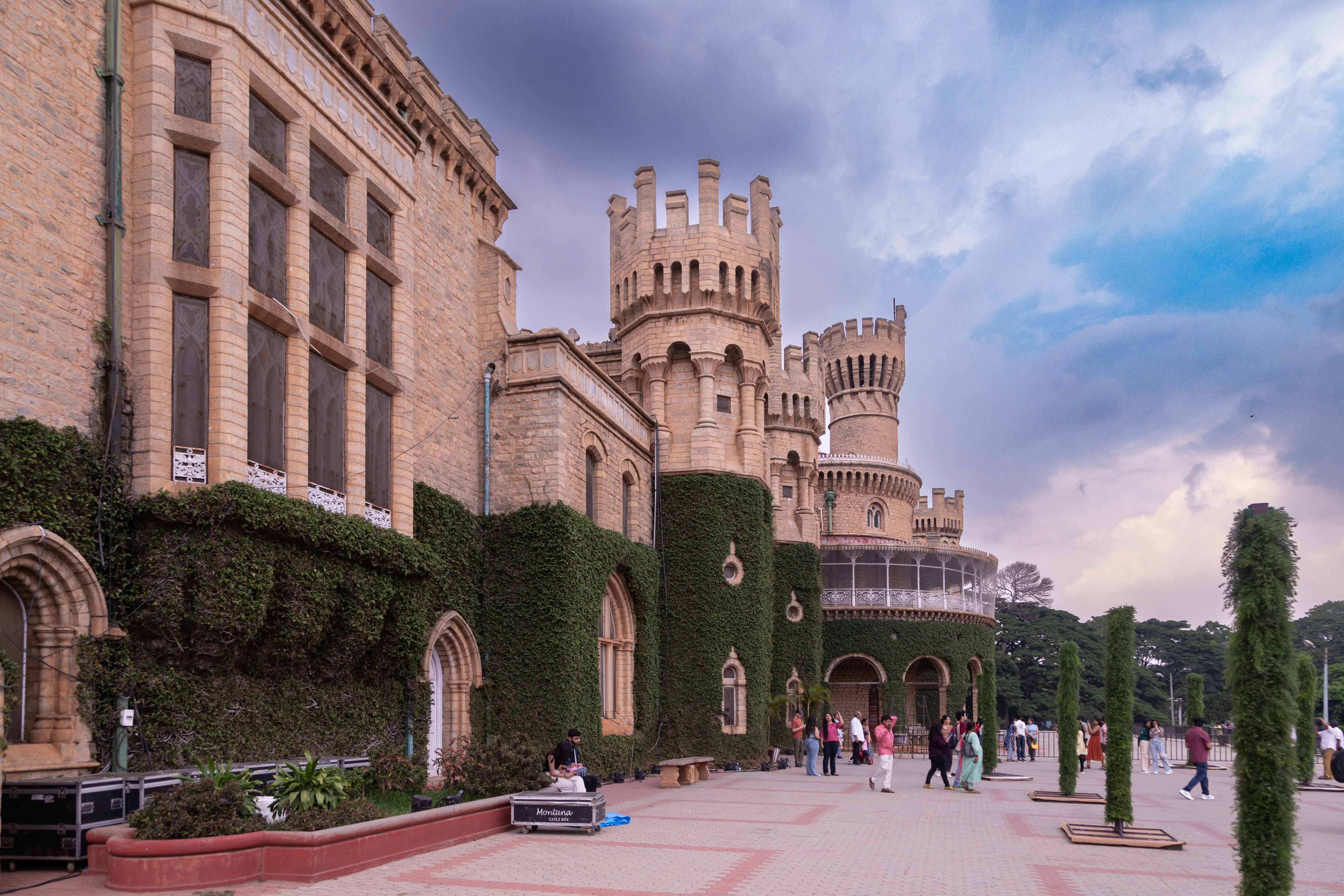 The majestic Bangalore Palace, a historical landmark in Bangalore, featuring grand stone architecture with ivy-covered walls and multiple towers. The palace courtyard is bustling with visitors and tourists, set against a backdrop of a partly cloudy sky. The scene captures the cultural and architectural significance of this iconic heritage site.