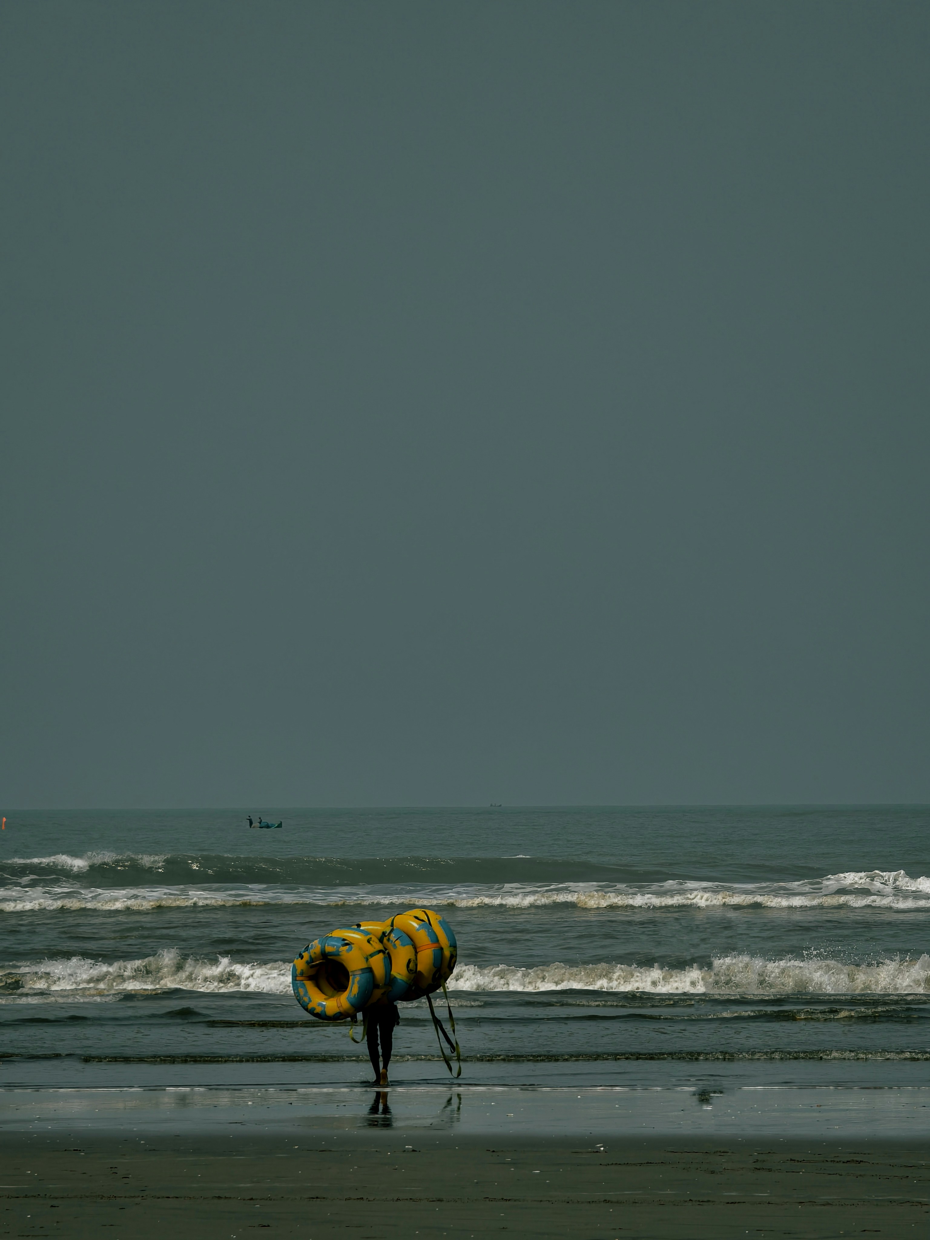 Bright yellow lifebuoy carried by a lone figure along a windswept beach, with gentle waves and a muted horizon.