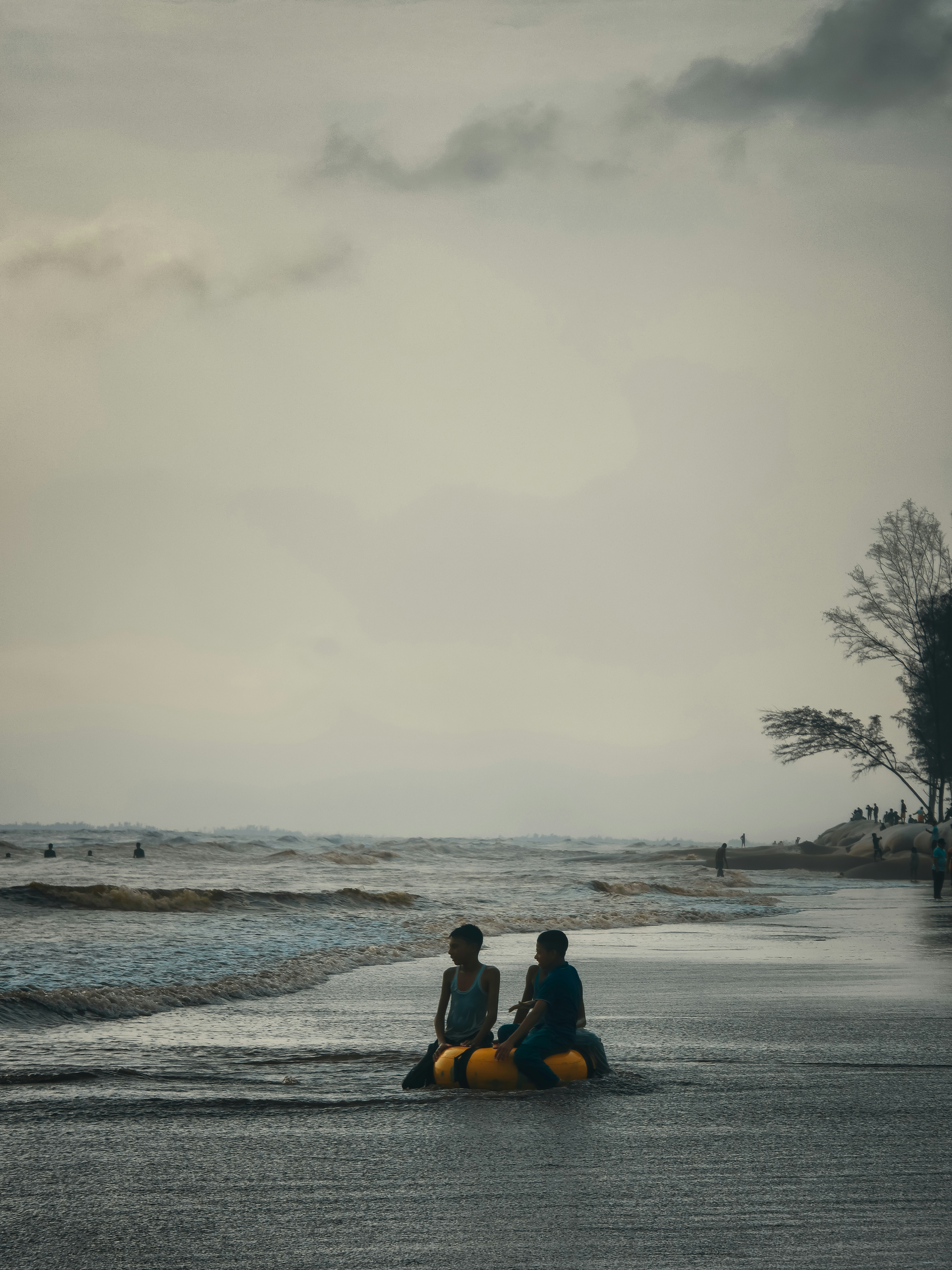 A photograph of two children riding a bright yellow inflatable tube along the calm shoreline at dusk, with gentle waves and a muted sky.