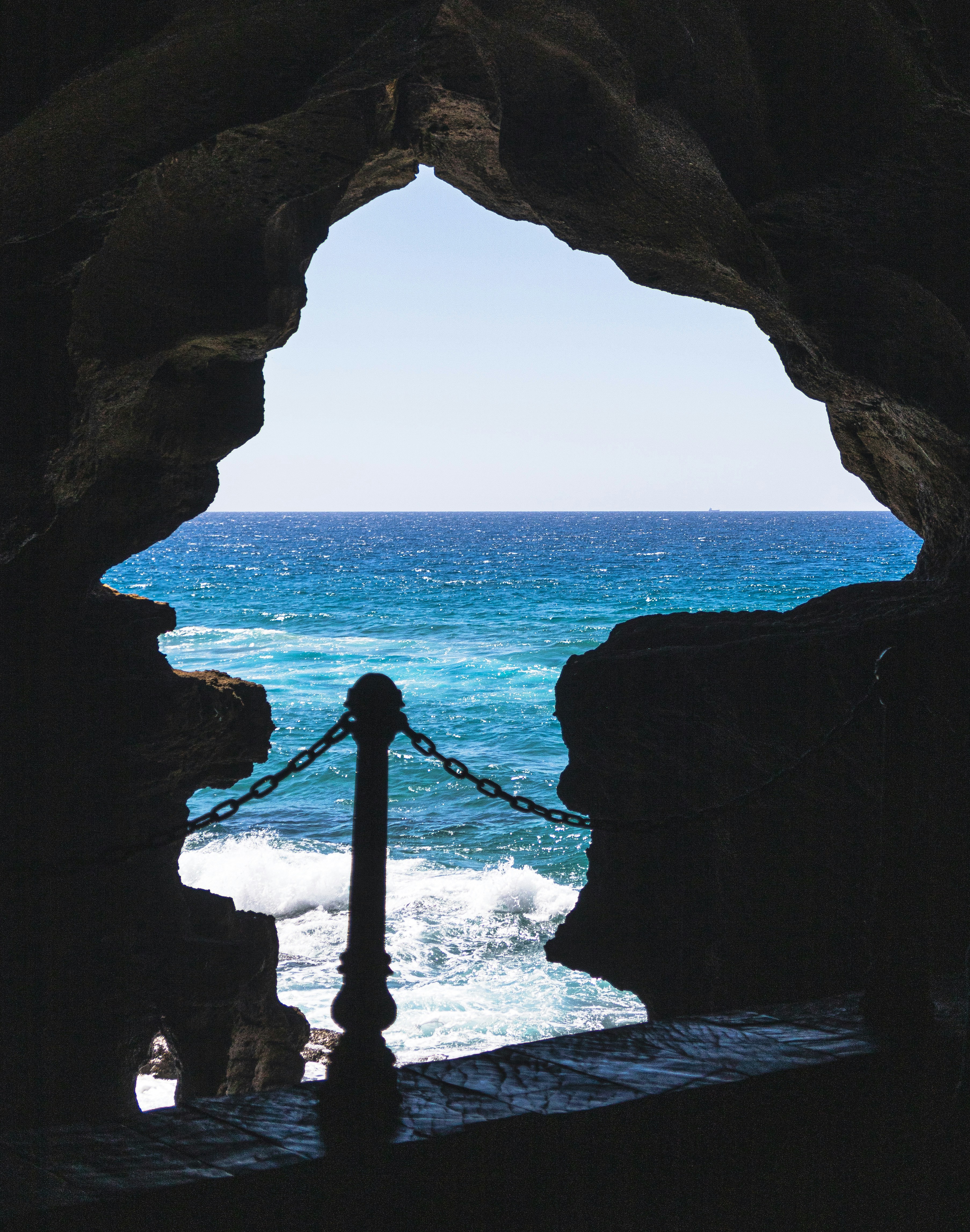 View of the ocean framed by a rocky archway, showcasing vibrant blue waters and white waves. A chain barrier adds an element of structure to the natural scene.