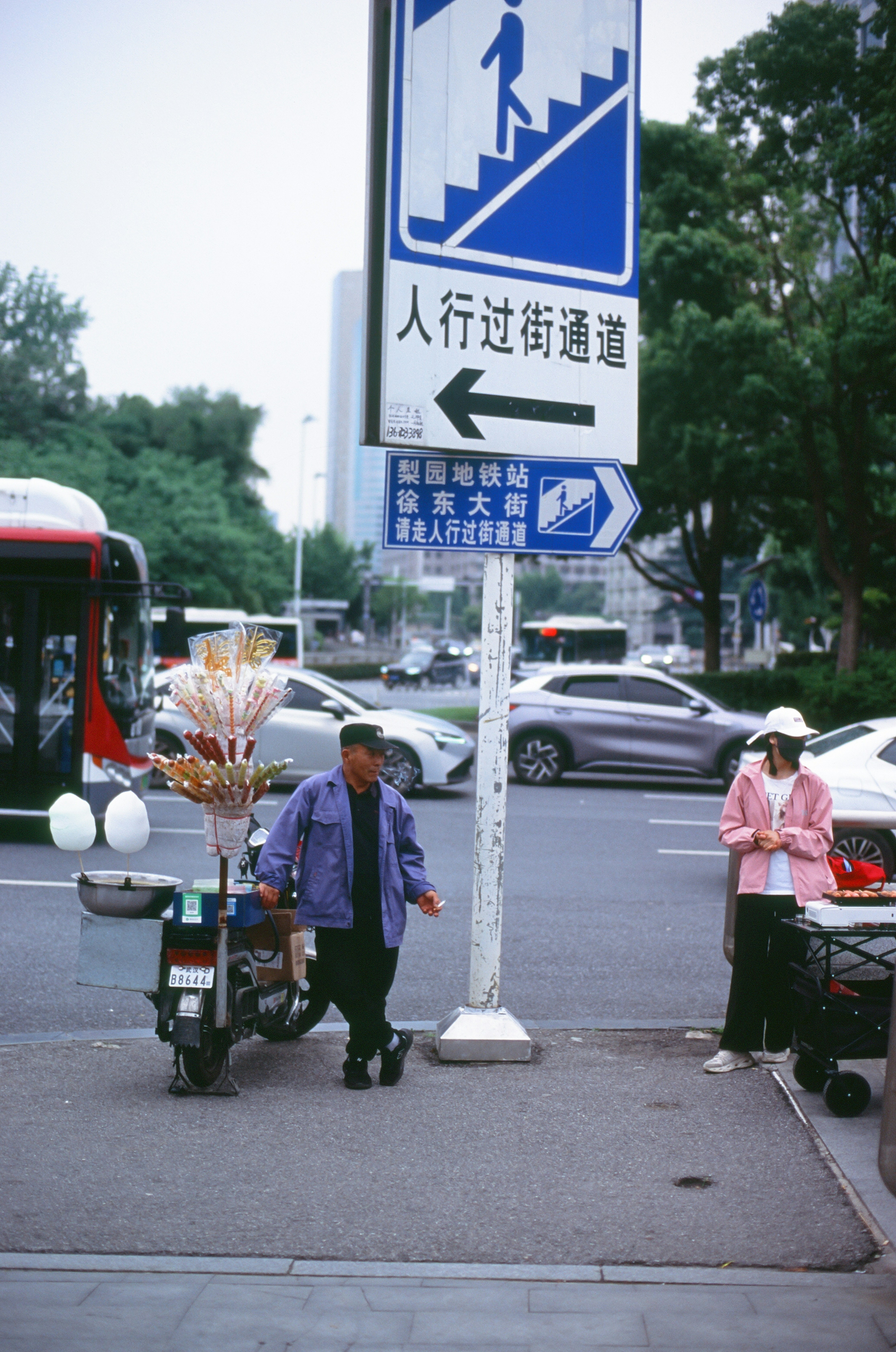 A street vendor in a purple jacket stands by a cart on a city sidewalk, framed by a tall blue traffic sign as pedestrians and cars pass in the background.