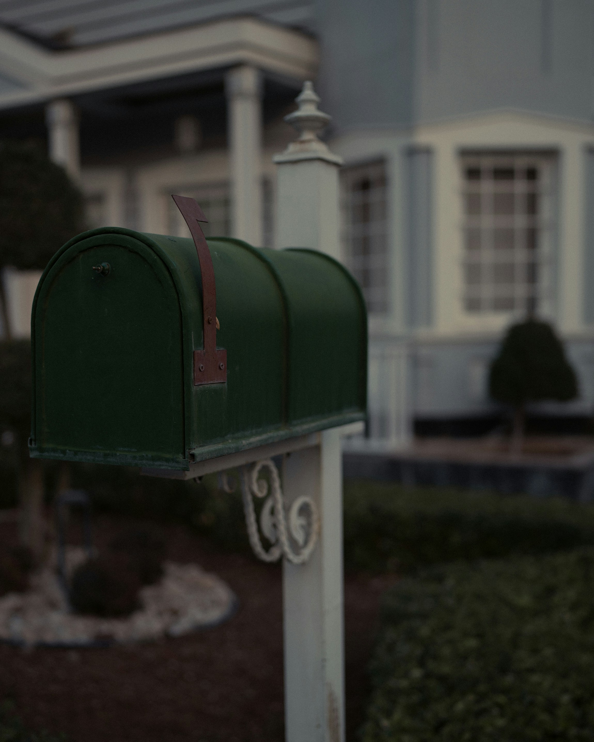 A vintage green mailbox stands prominently on a white post, surrounded by neatly trimmed hedges and a serene home backdrop. The scene evokes a sense of nostalgia and tranquility.