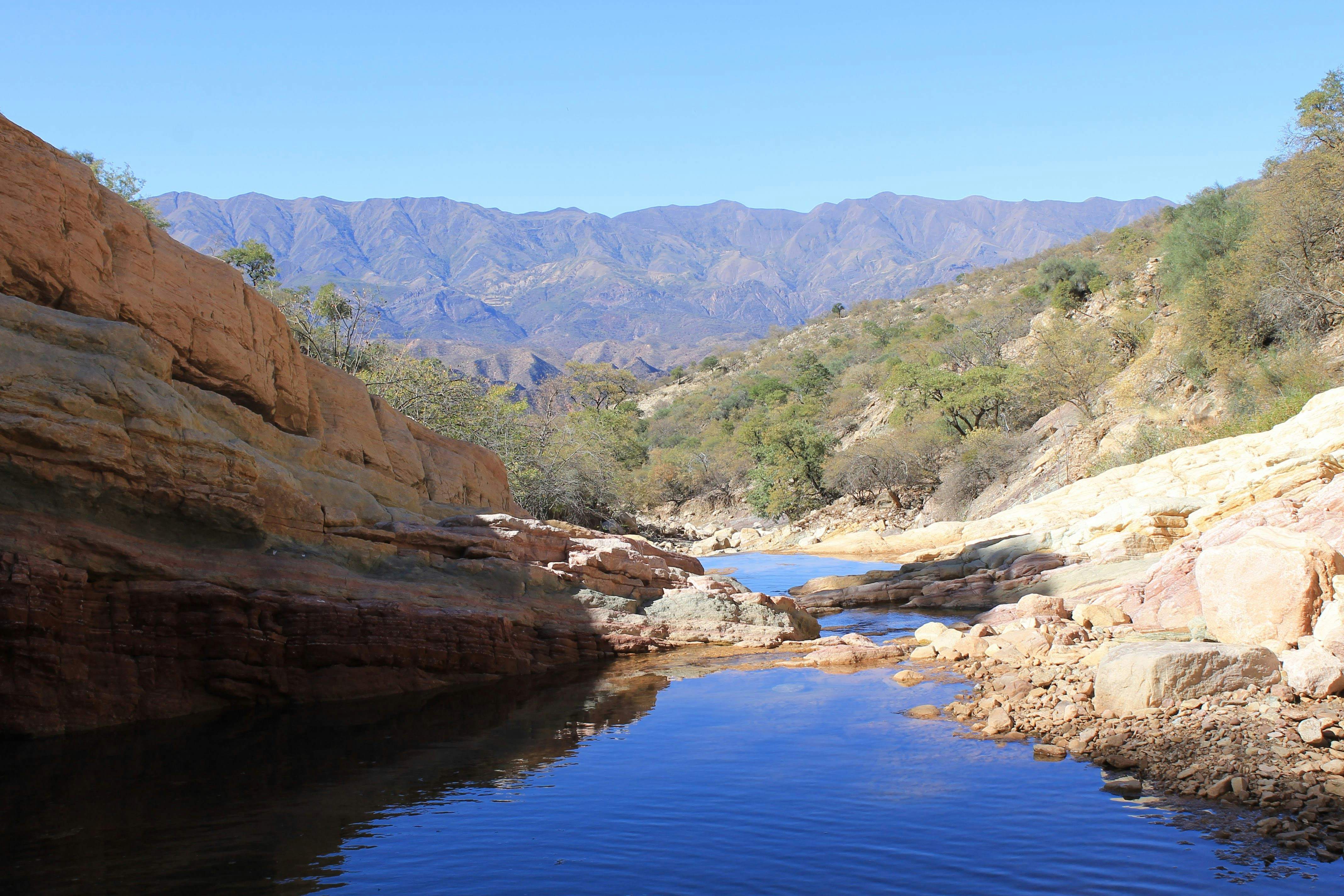 Clear blue river winding through rocky canyon with distant mountain backdrop under a bright sky.