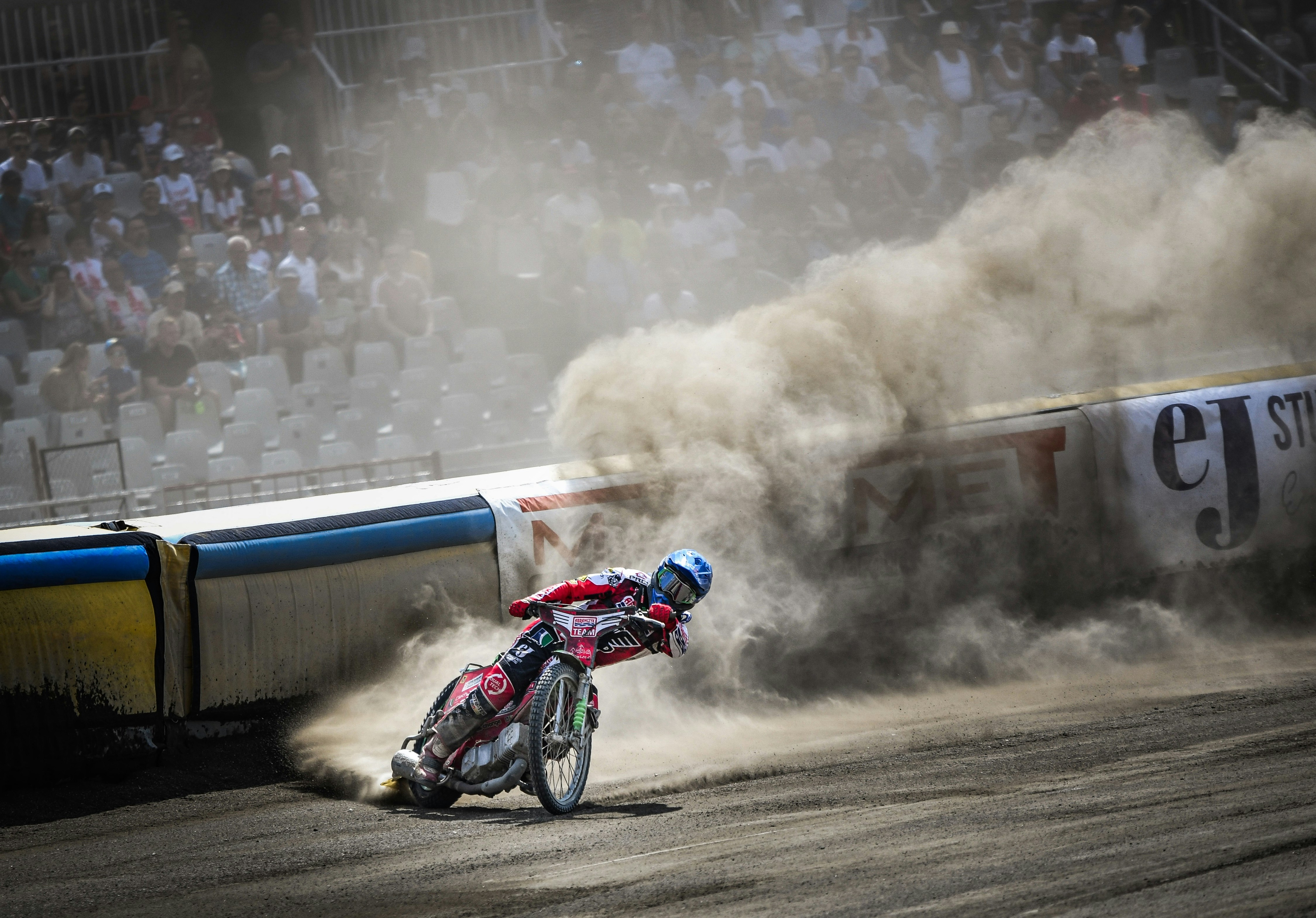 Motorcyclist leaning into a sharp track curve, trailing a cloud of dust with blurred spectators in the background.
