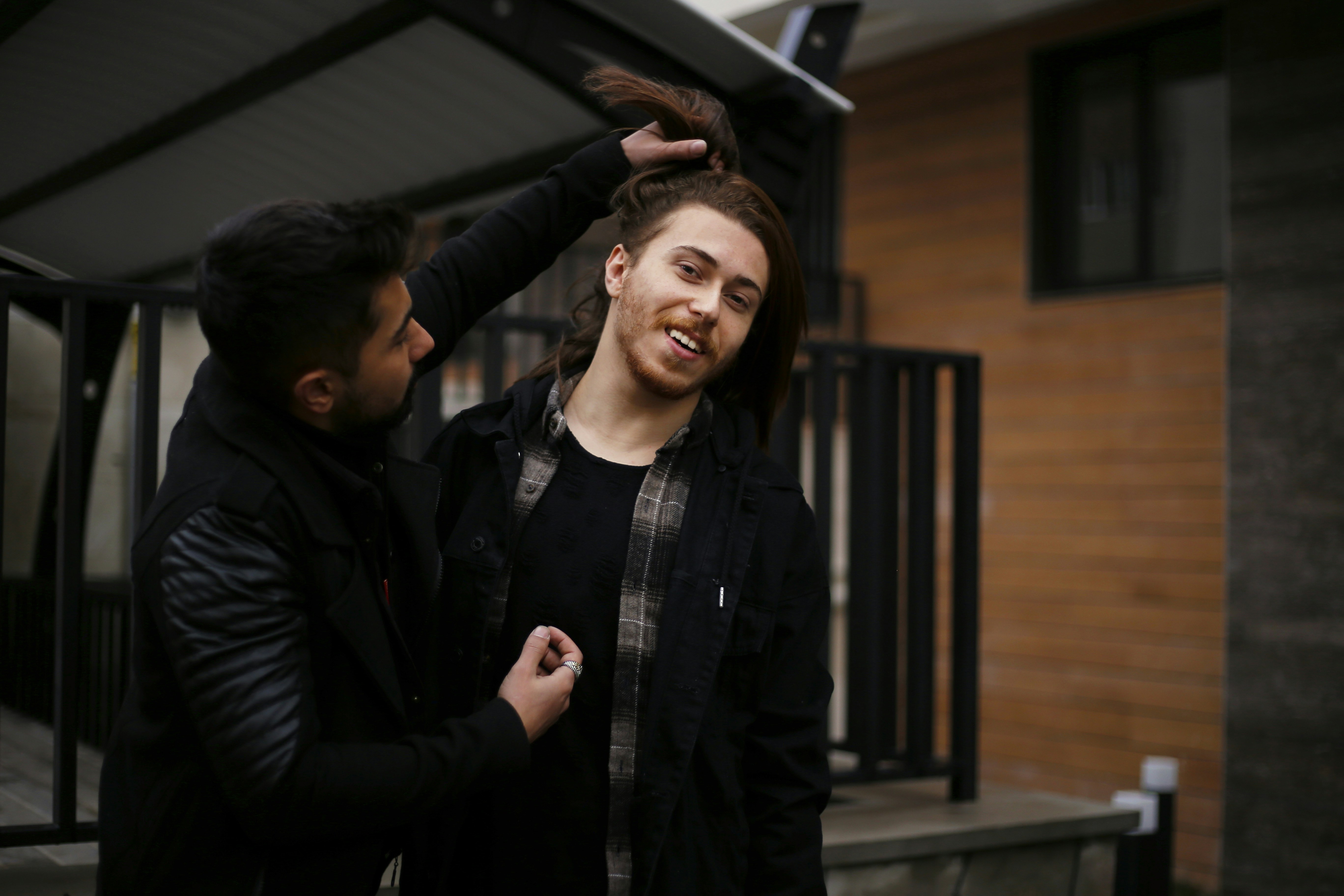 Two men on a wooden porch; one adjusts the other's hair as they share a candid moment during an outdoor shoot.