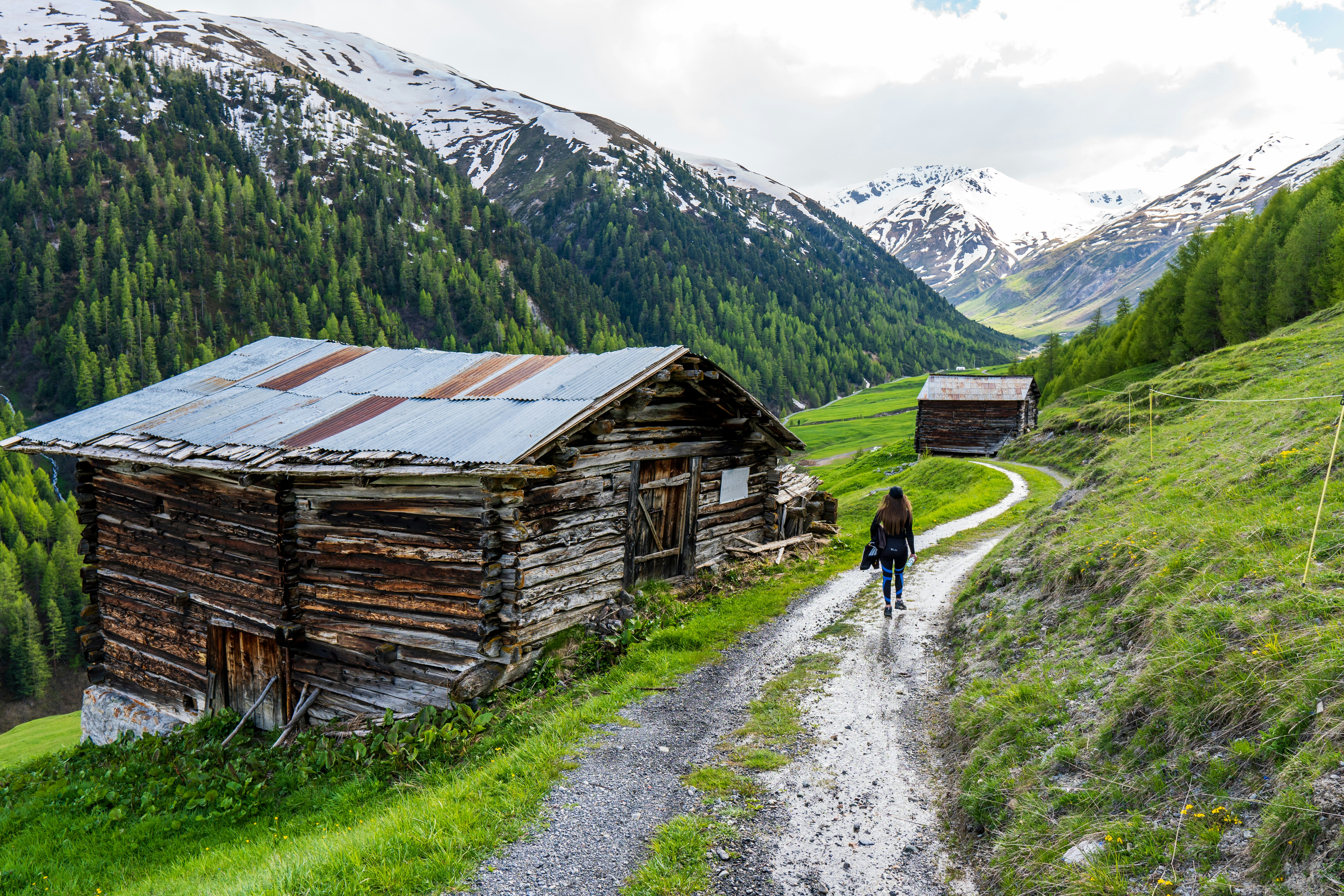 Hiker on a Weathered Alpine Path Between Rustic Cabins and Snowy Peaks