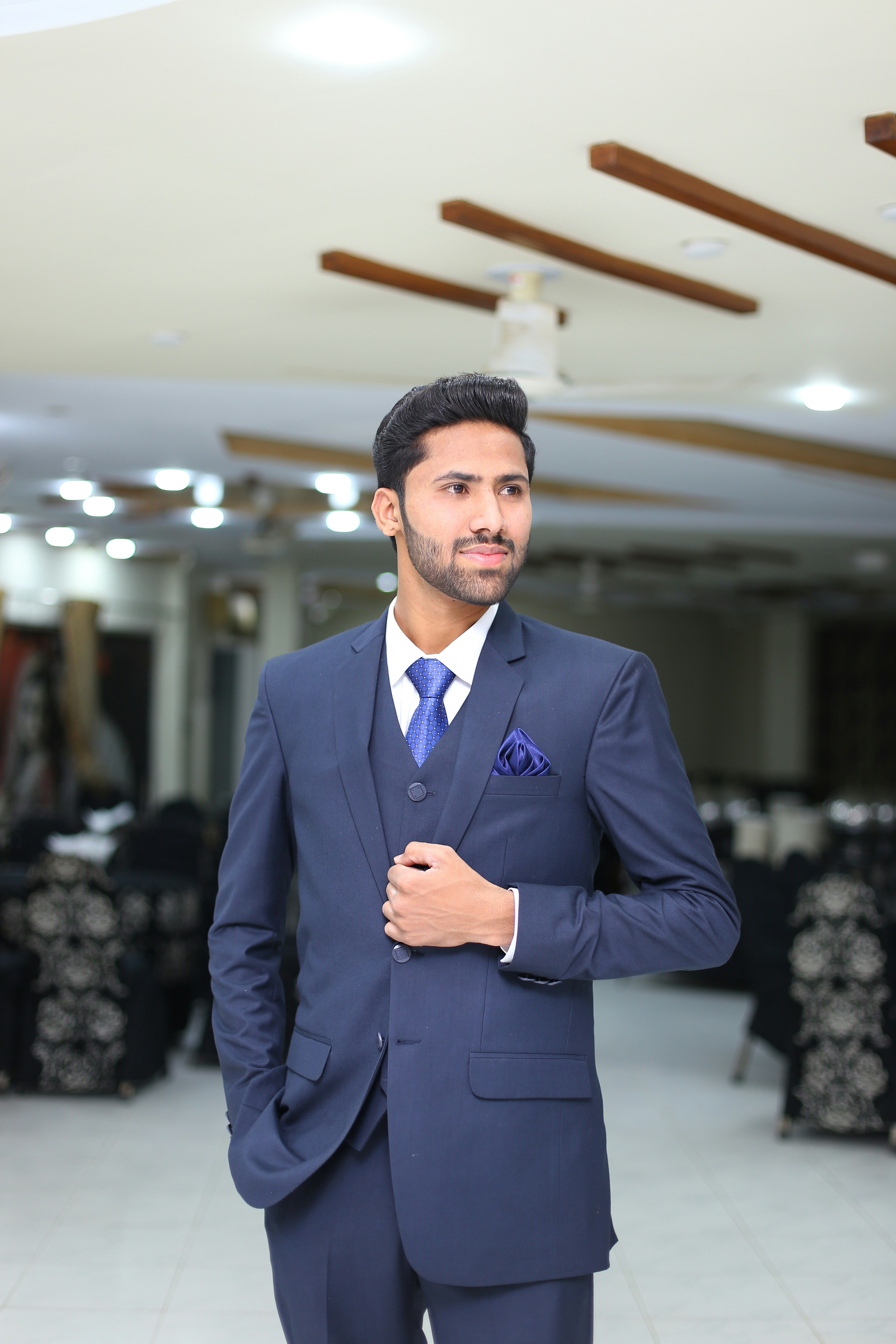 A well-dressed man in a suit poses confidently in a banquet hall, showcasing a blend of style and poise. His thoughtful expression adds depth to the composition.