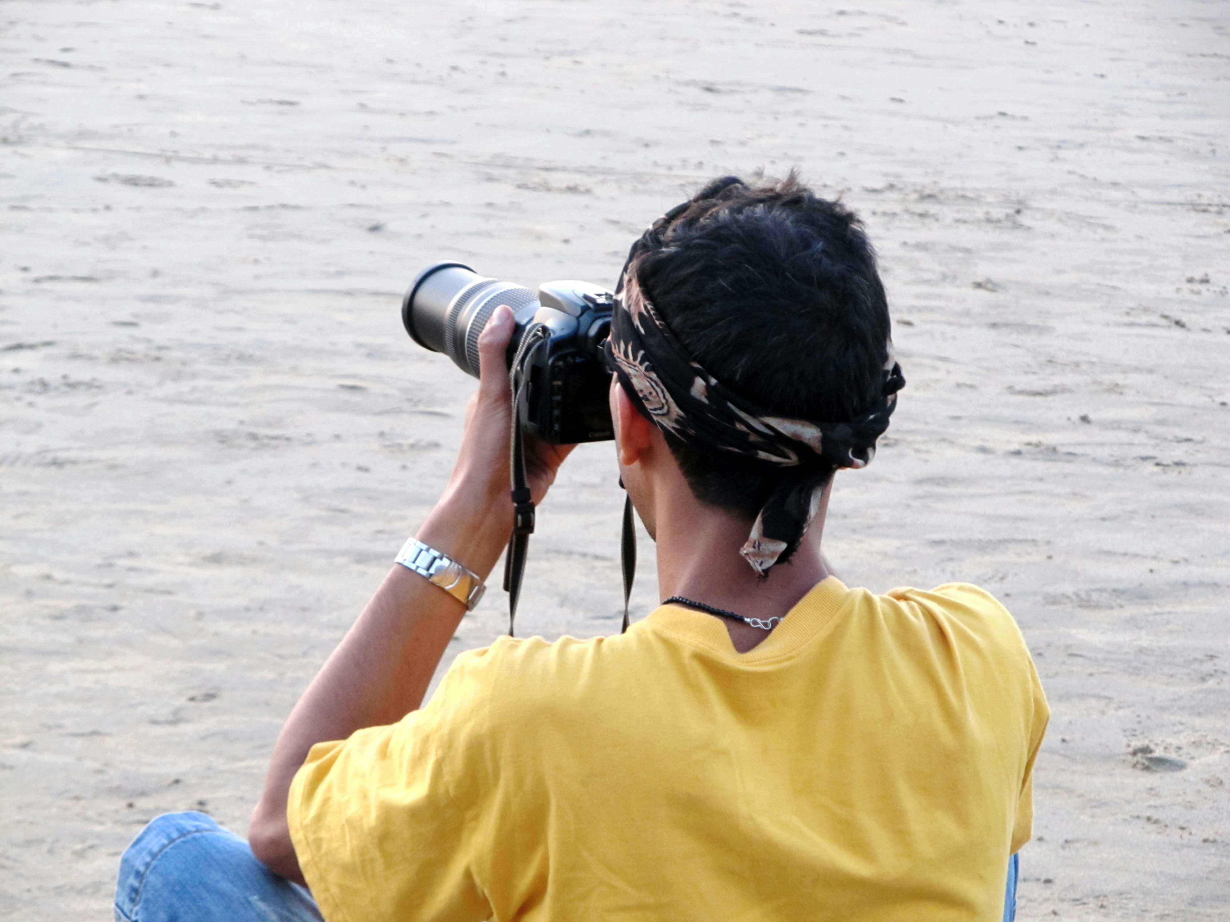 Photographer in a yellow shirt aims a camera with a long lens at a sandy beach.