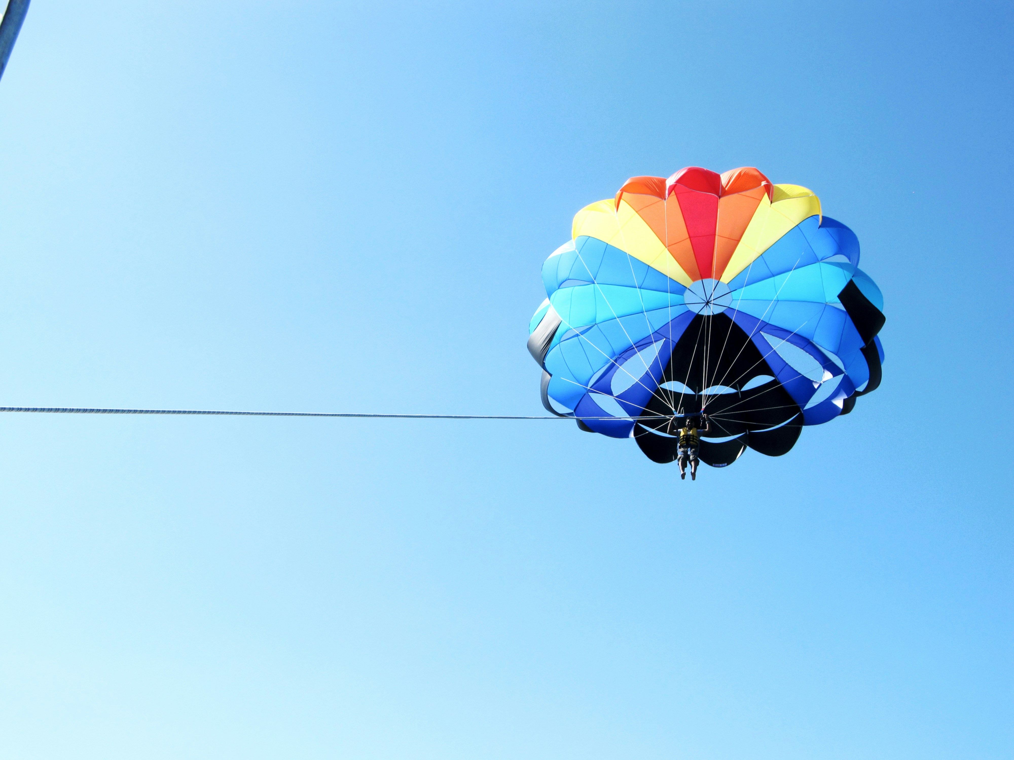Colorful paraglider soars beneath a rainbow canopy across a crisp blue sky, with a single utility line cutting through the scene. A lone rider hangs beneath the vibrant canopy, framed by bright daylight.