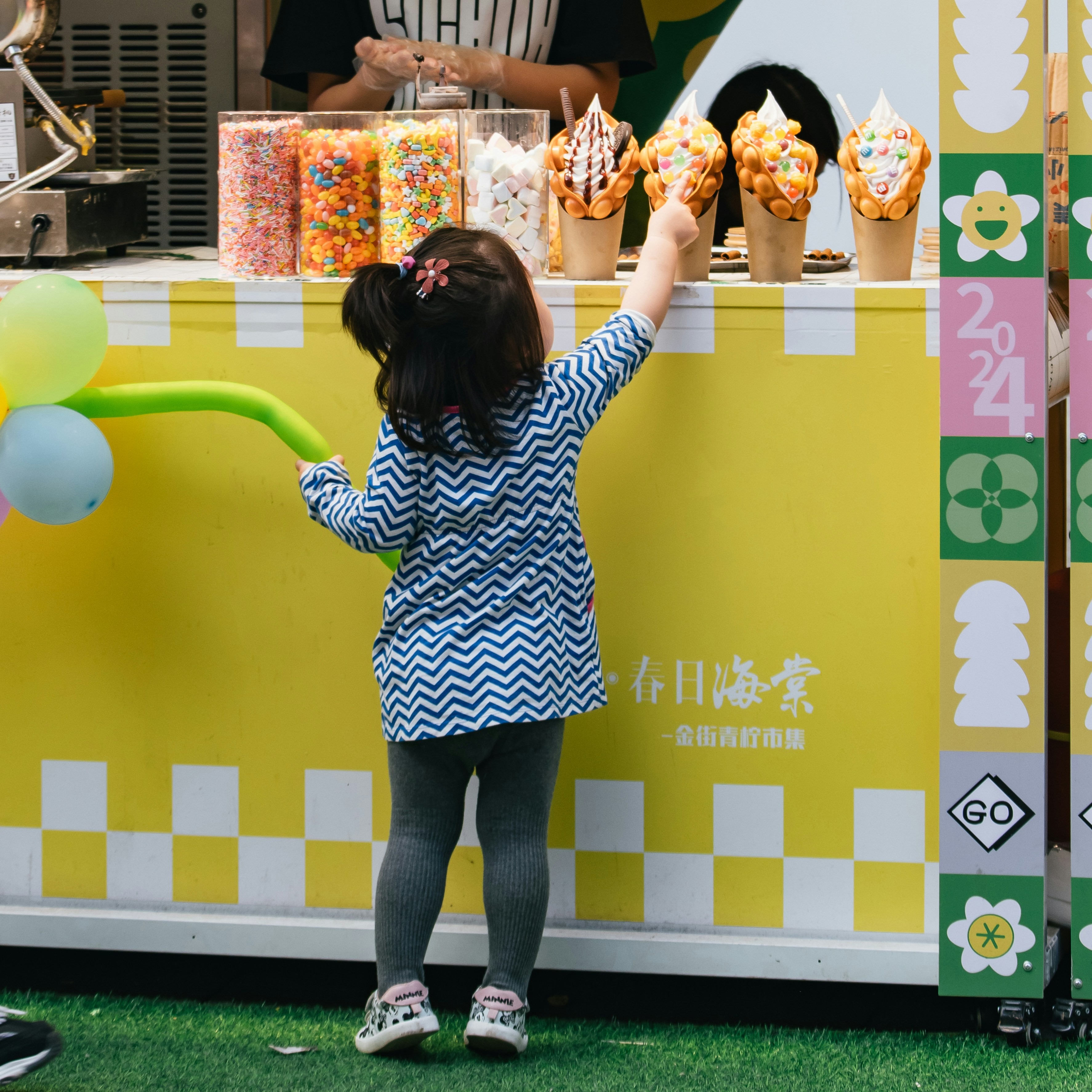 A young girl in a blue zigzag dress reaches toward oversized ice-cream cones at a bright yellow carnival stall, with candy jars and a vendor visible in the background.