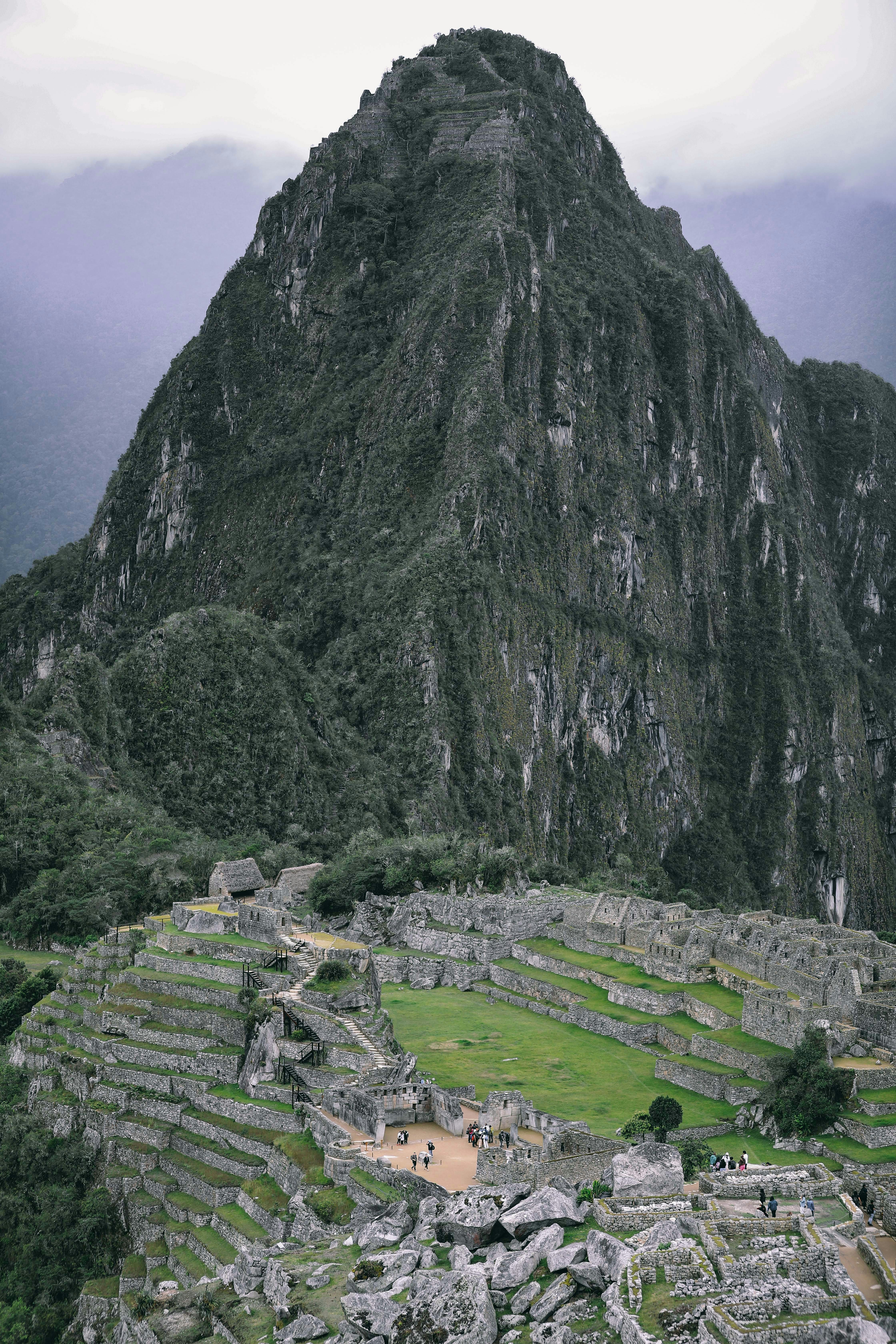 Aerial view of Machu Picchu, showcasing its intricate stone terraces and structures against a backdrop of steep, lush mountains. Visitors explore the historic site.