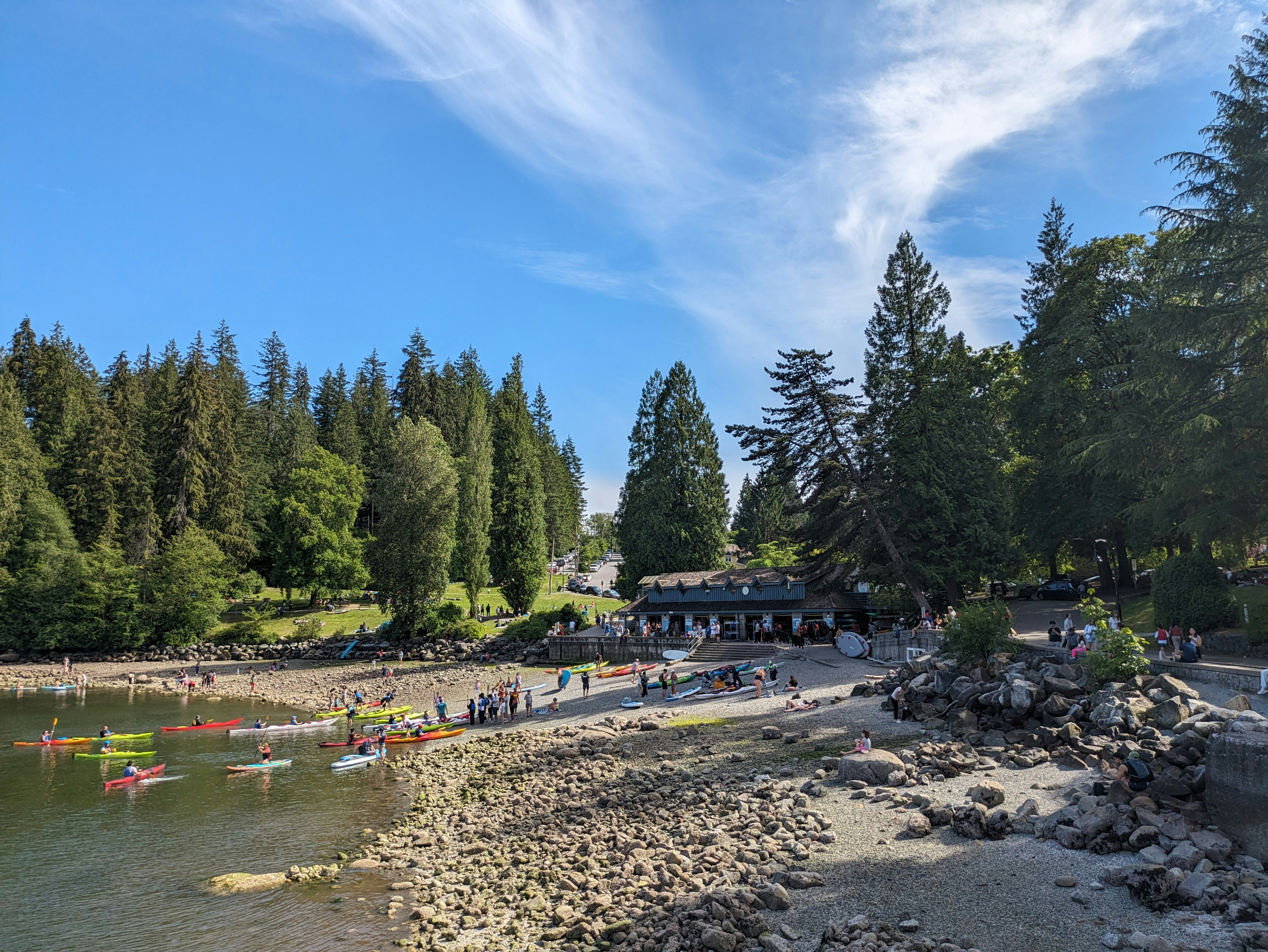 Photograph of a sunlit lakeside park with a pebble beach, colorful kayaks along the water, and a rustic building among tall pines. People enjoy the scene along the shore under a bright blue sky.