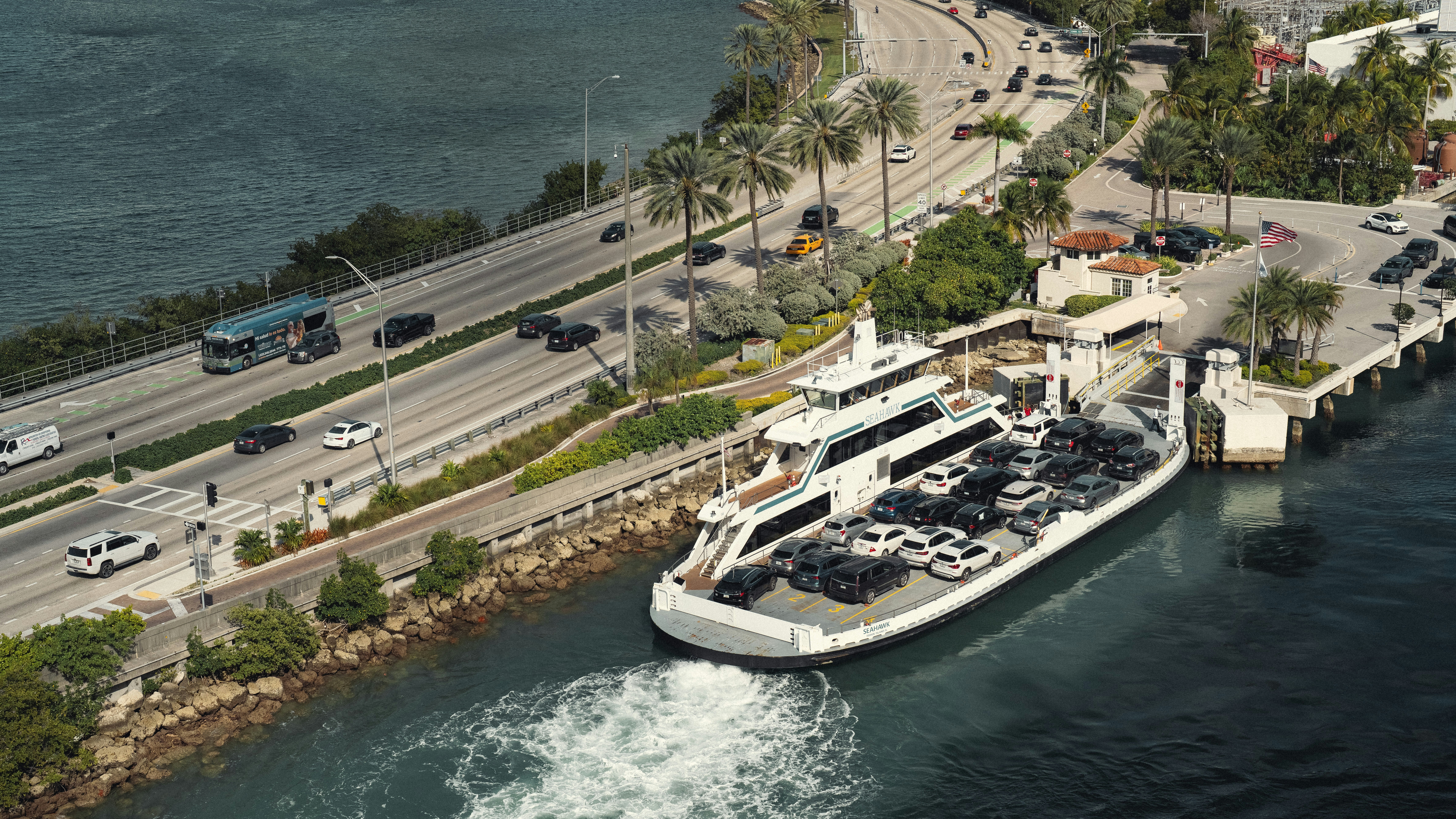 This aerial shot shows a car ferry loaded with vehicles departing from a dock in Miami. The ferry navigates the calm waters alongside a palm tree-lined road bustling with traffic. This scene highlights the intersection of urban infrastructure and maritime travel, capturing the essence of island life and transportation in a coastal city setting.