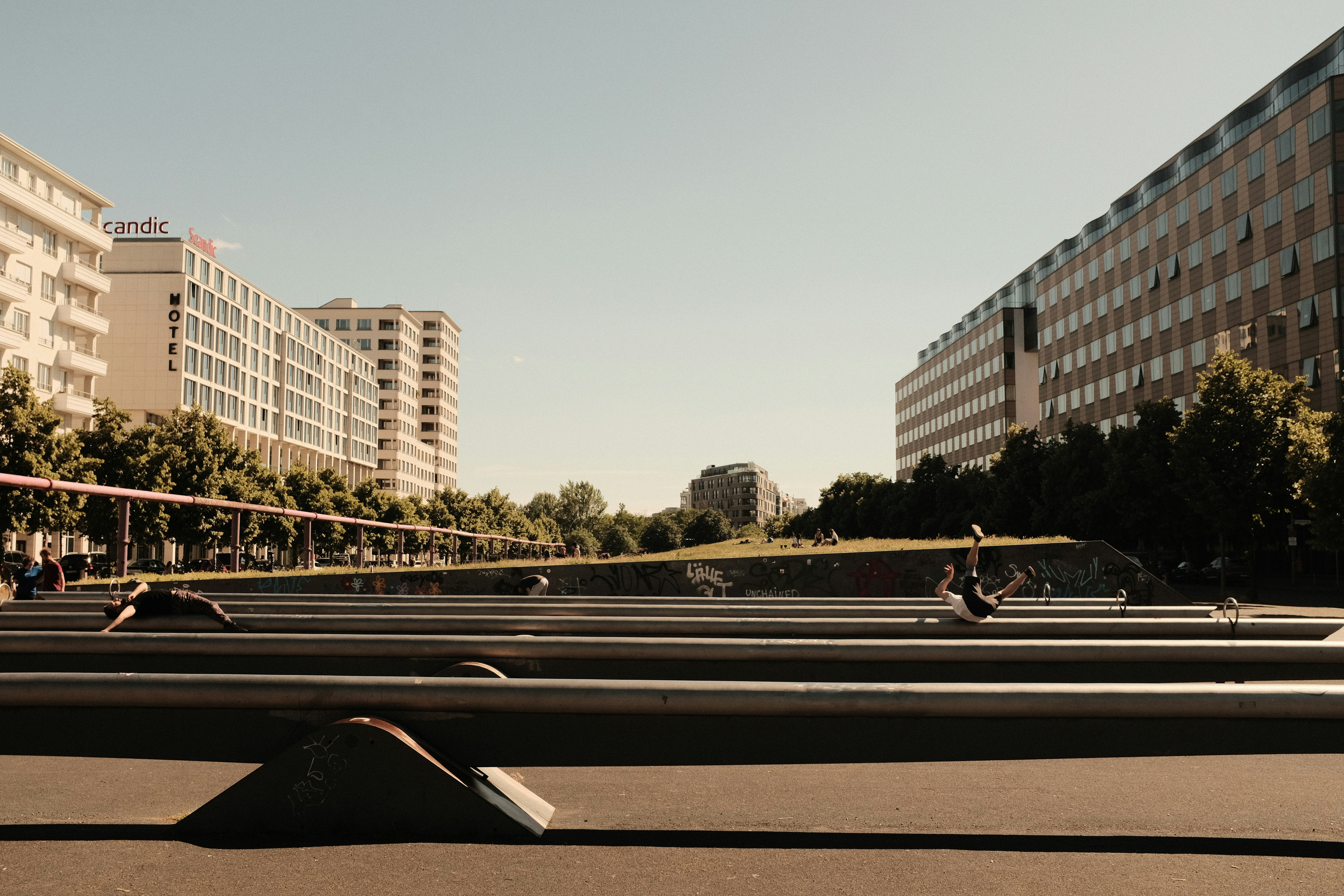 Skaters performing tricks on metal rails in a vibrant urban setting, framed by modern architecture and lush greenery.