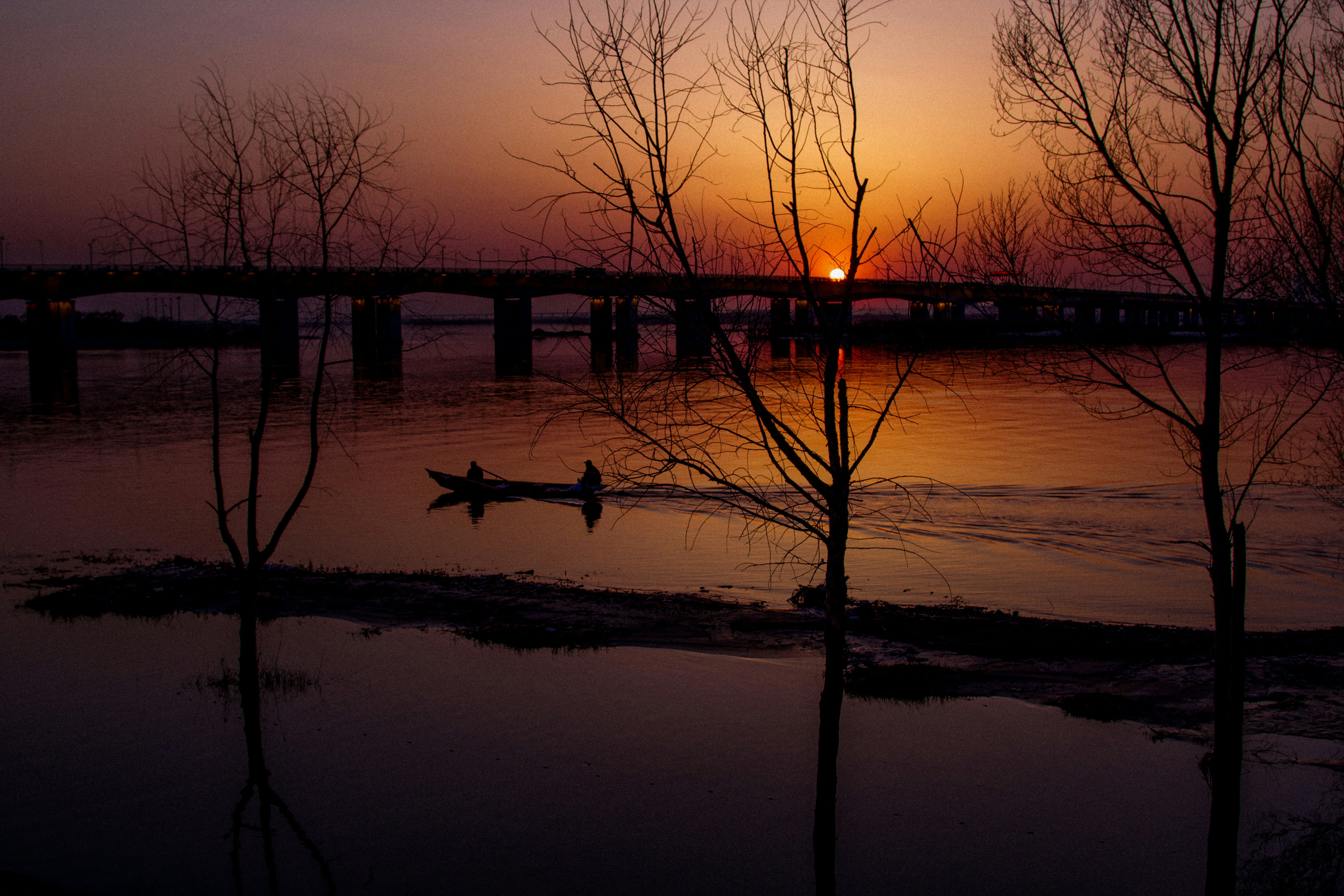 A lone fisherman navigates a tranquil river at sunset, silhouetted against a vibrant sky. Bare trees frame the scene, enhancing the serene atmosphere.