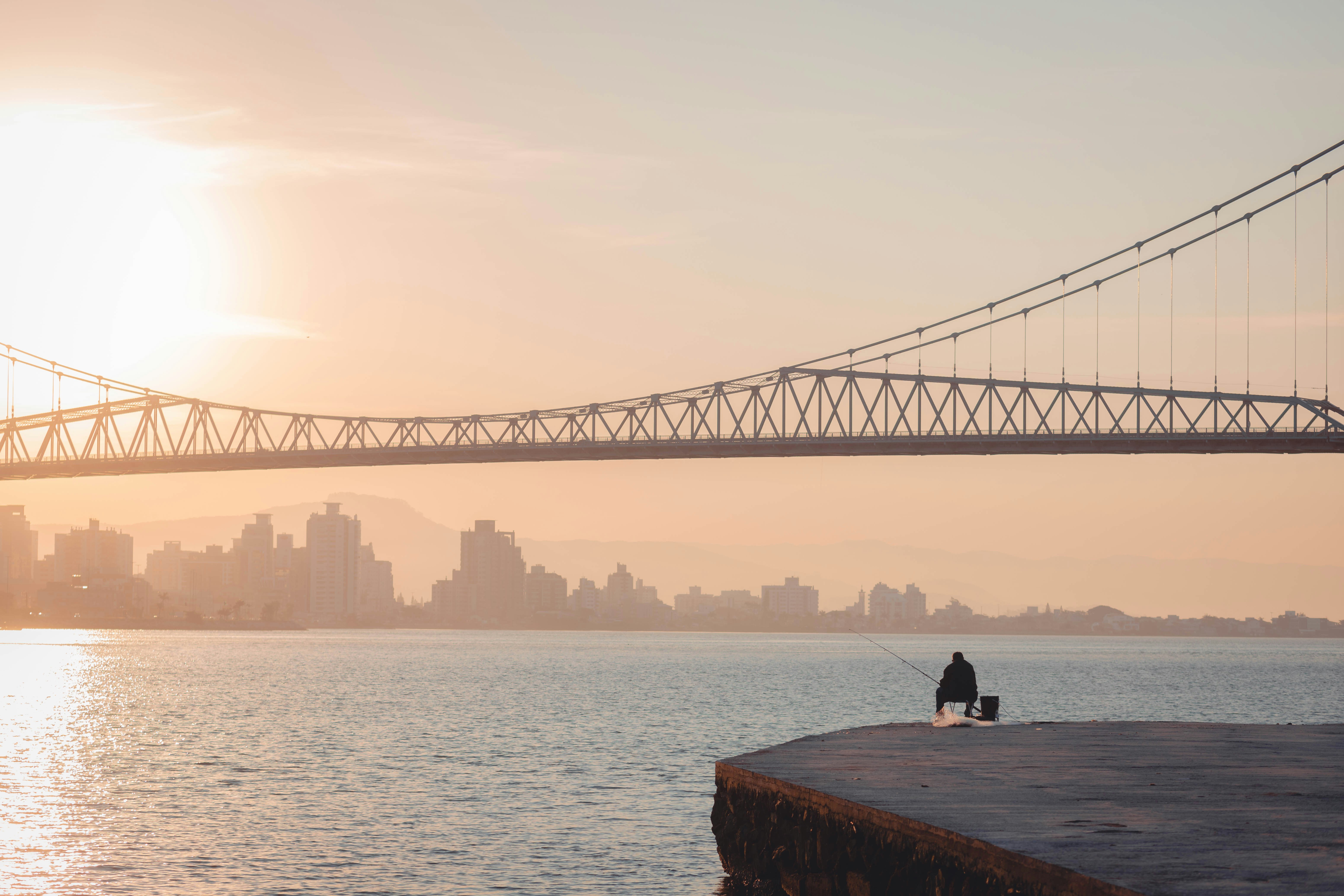 Edge of Quiet Light: lone figure on the harbor pier beneath a waking bridge