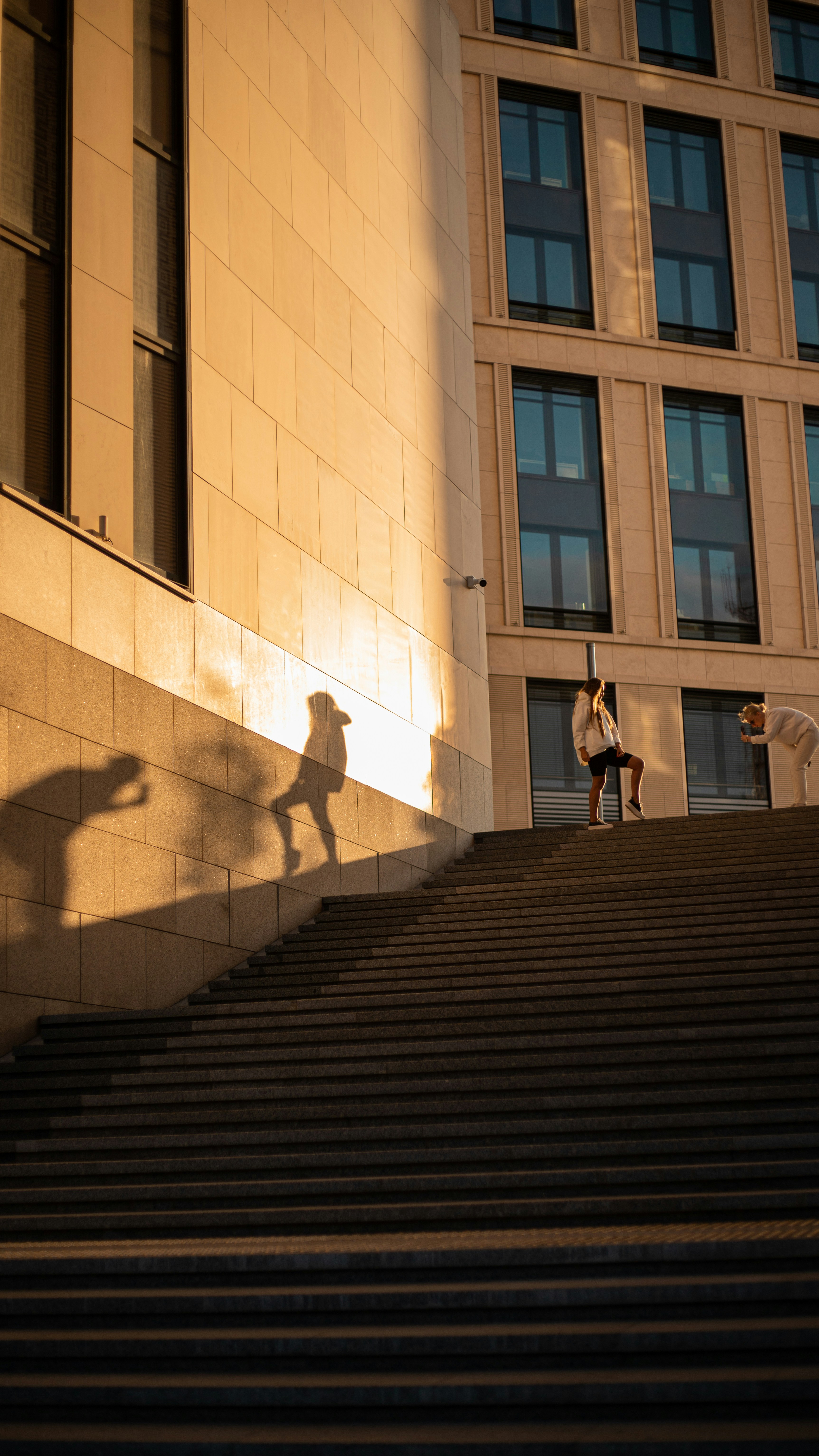 Two figures interact on a staircase, their shadows creating a dynamic composition against a sunlit wall. The play of light adds depth to the scene.