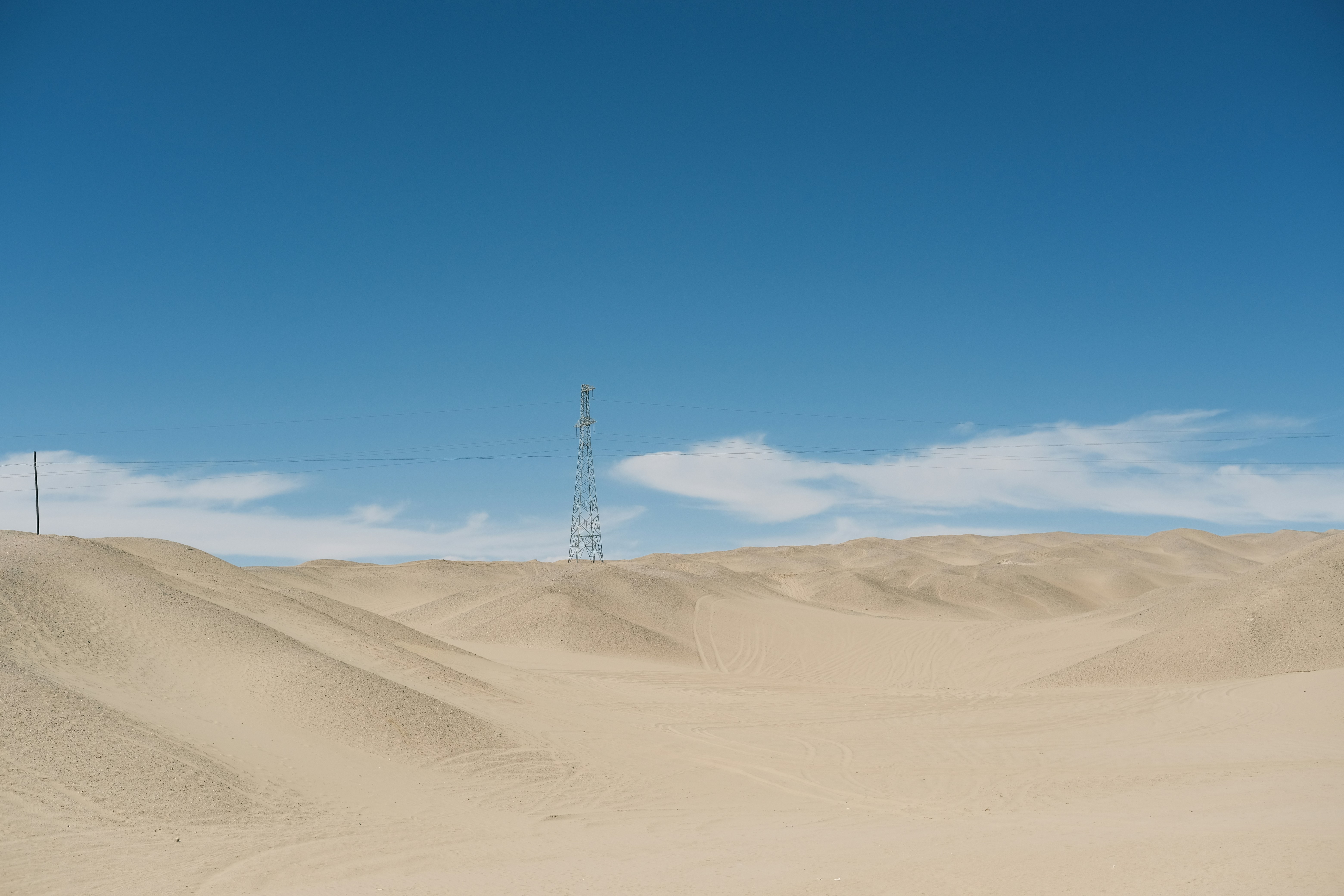 The image captures the essence of solitude in a vast desert landscape. Against the backdrop of an azure sky, a solitary utility pole stands resolute, its power lines stretching across the sandy terrain. The scene evokes a sense of isolation, where nature’s canvas unfolds uninterrupted, punctuated only by sparse vegetation and low-lying hills. Here, the stark contrast between the expansive desert and humanity’s minimal imprint invites contemplation.