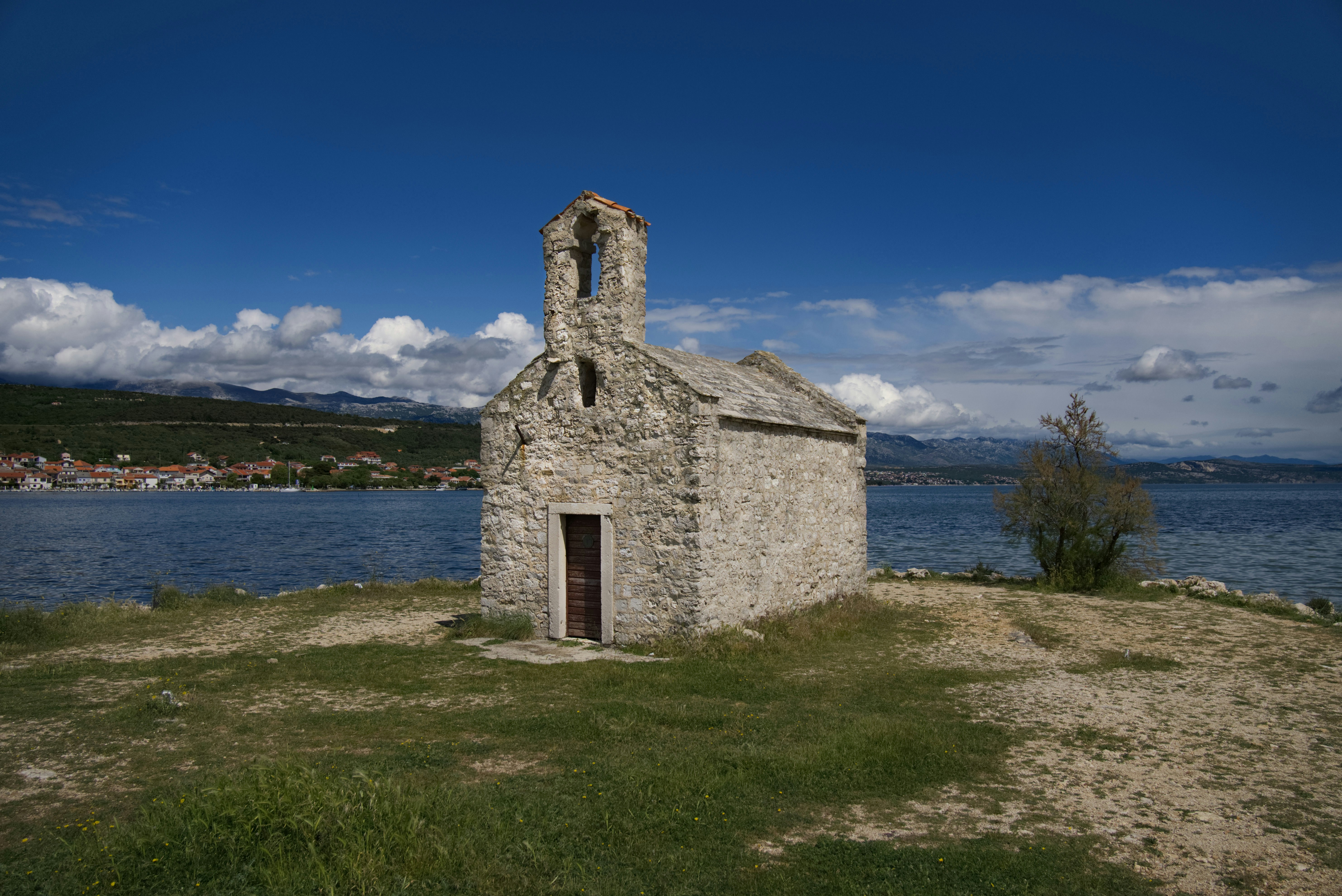 Ruined chapel by the lake: stone walls, blue sky, and distant village