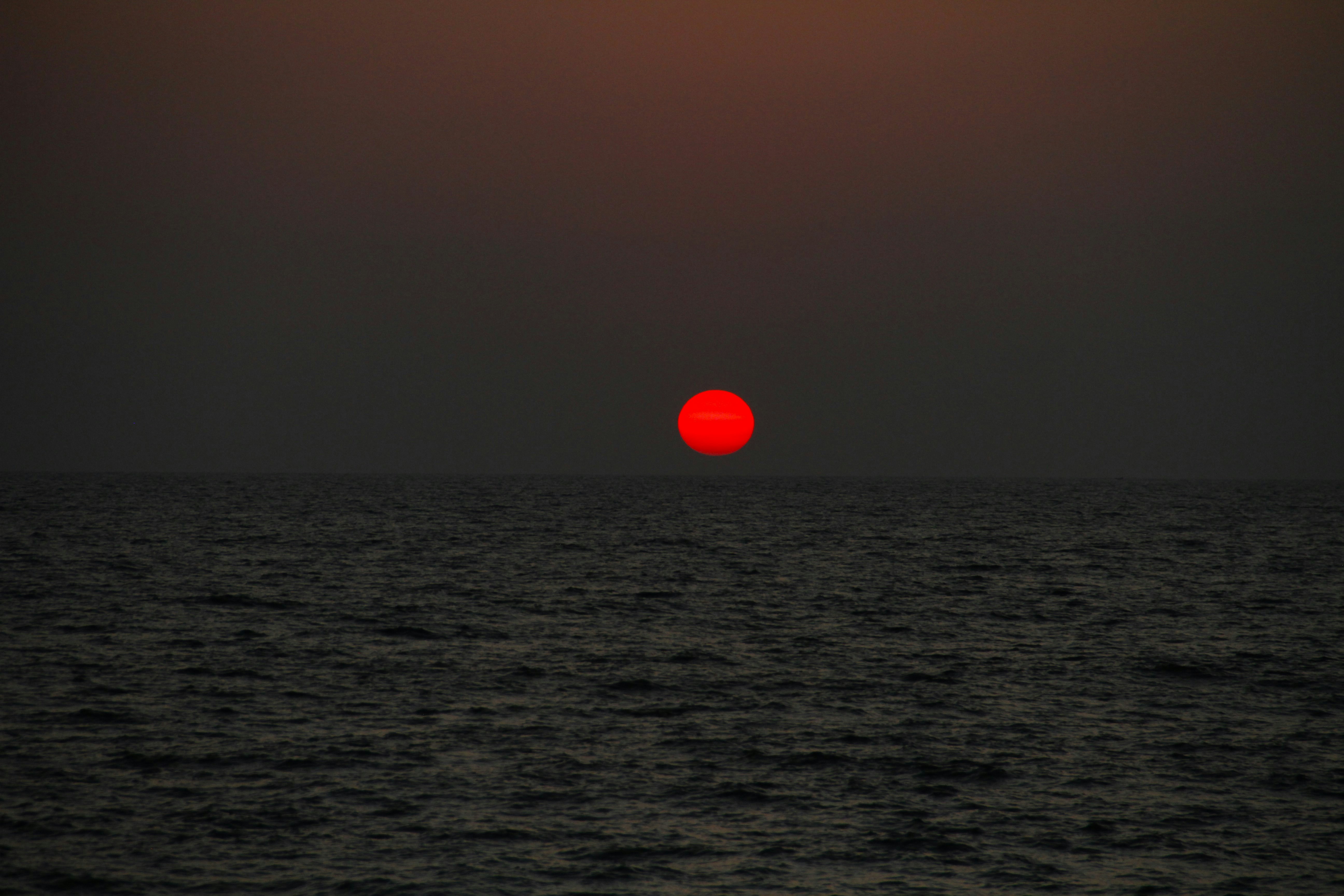 Vibrant red sun setting over a calm ocean, casting reflections on the water's surface.